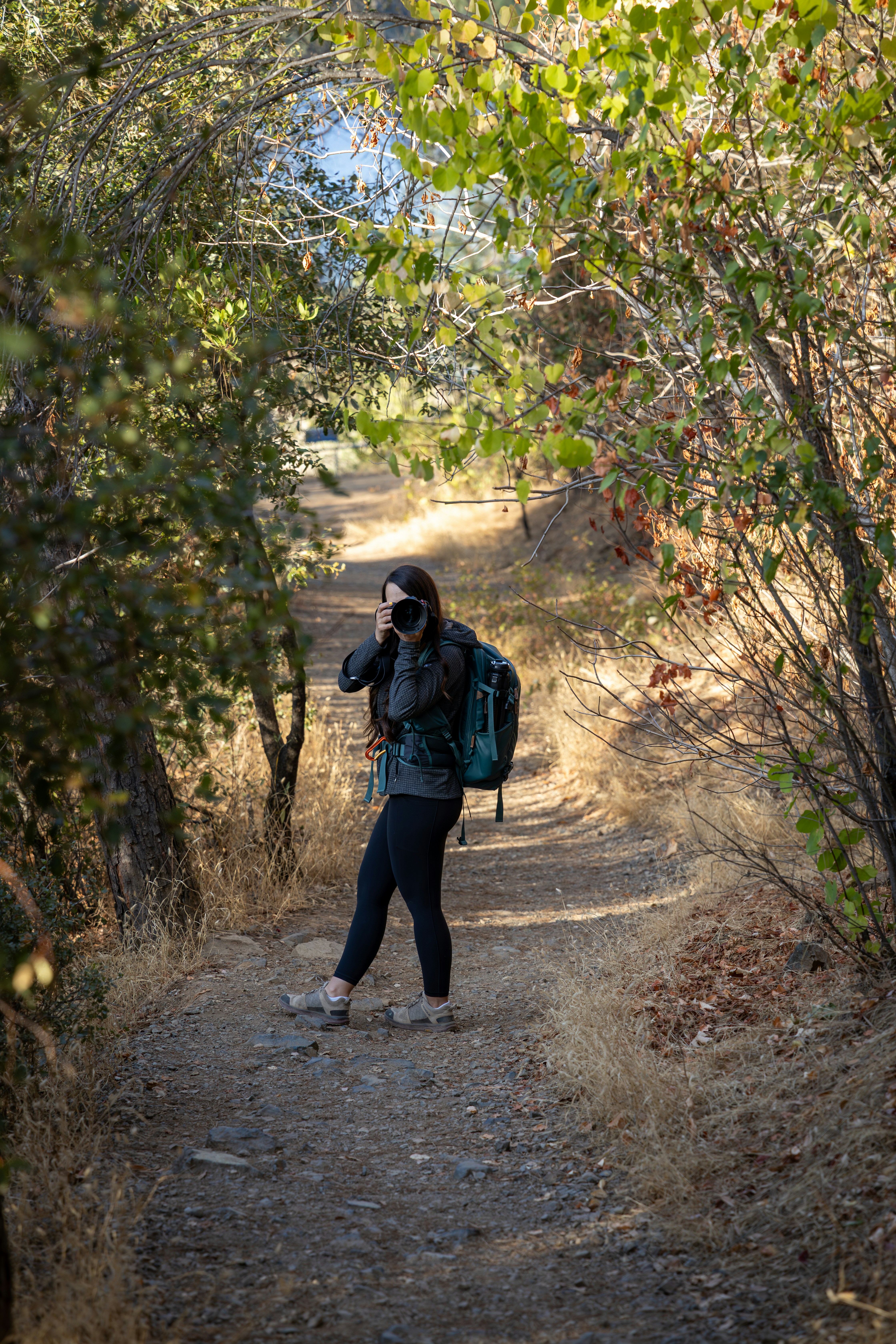 Hiker with a camera stands on a winding trail surrounded by lush greenery and autumn foliage.