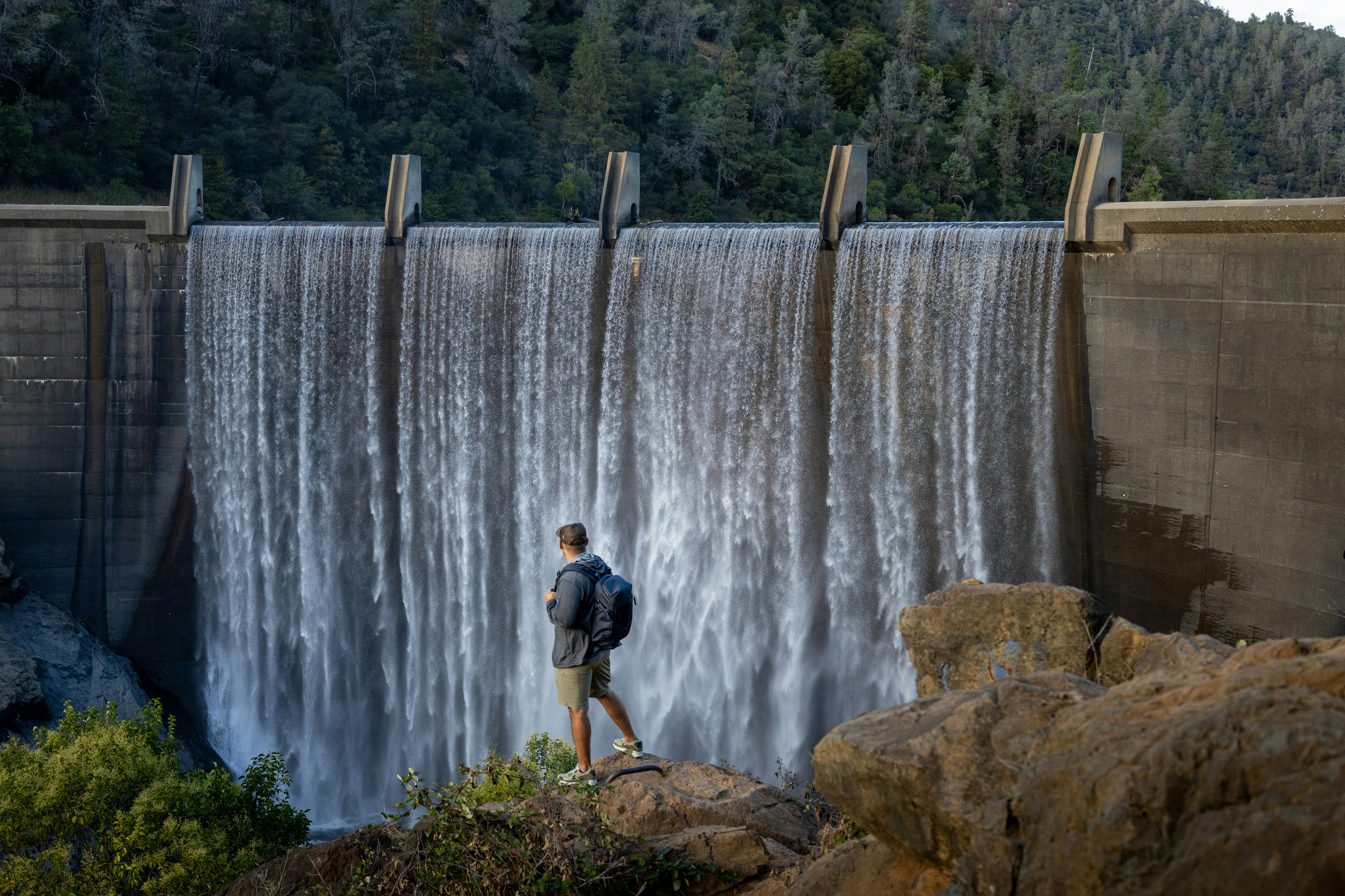 Hiker at the Lip of a Cascading Spillway Beneath a Forested Dam