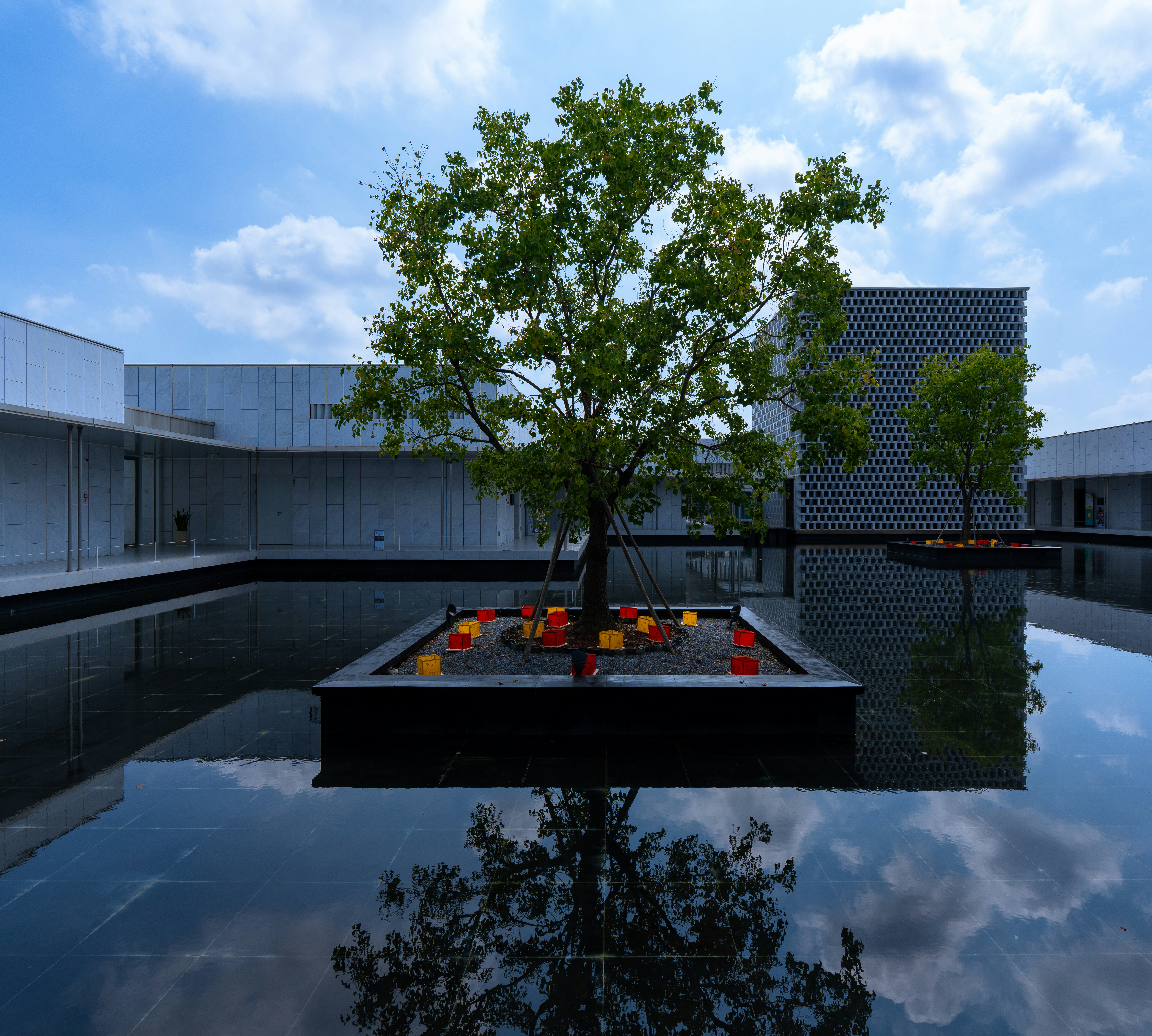 A serene courtyard featuring a central tree surrounded by colorful stones, mirrored in a still pond under a cloudy sky.