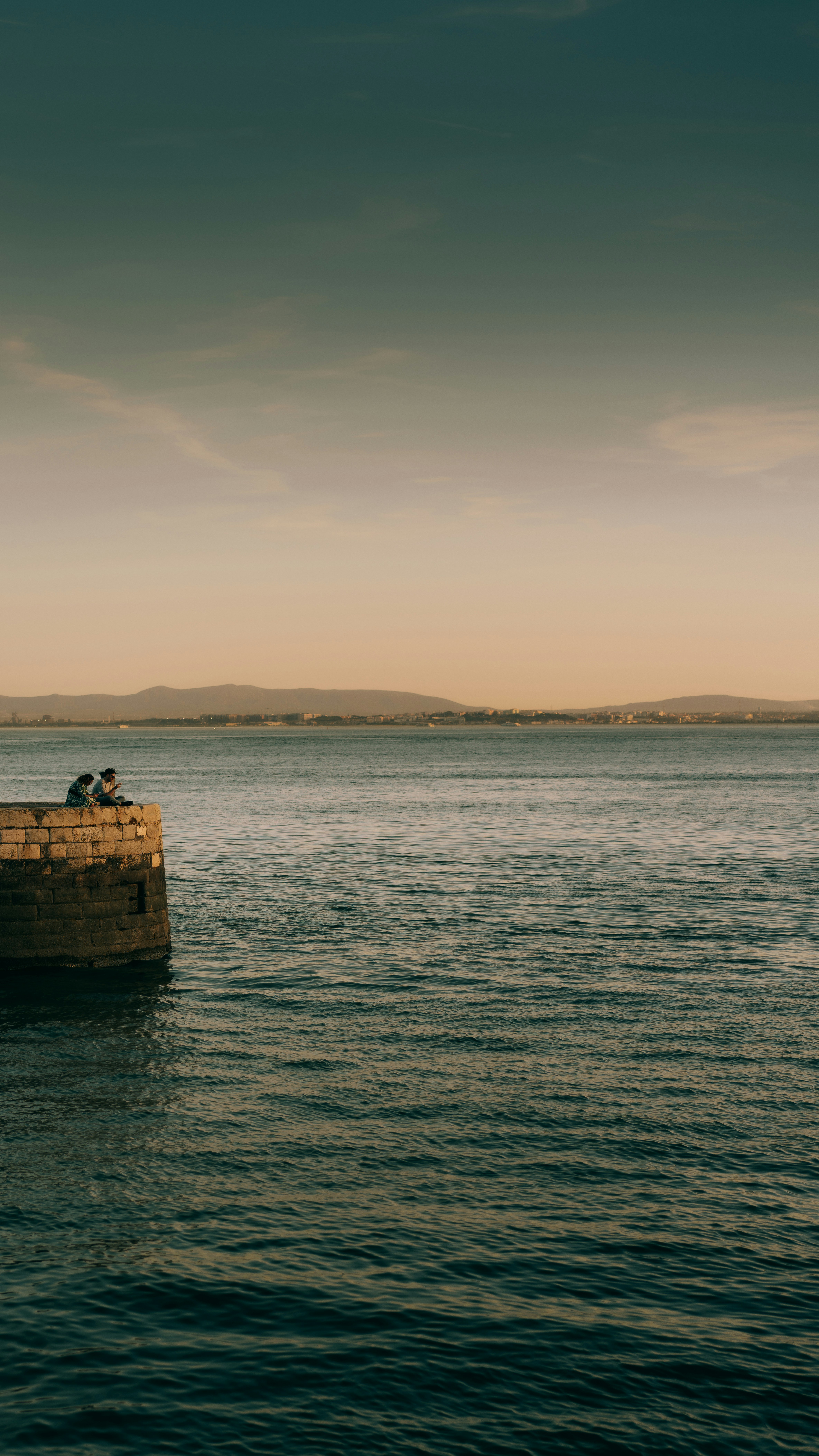 Two people sit on a stone pier overlooking the ocean.