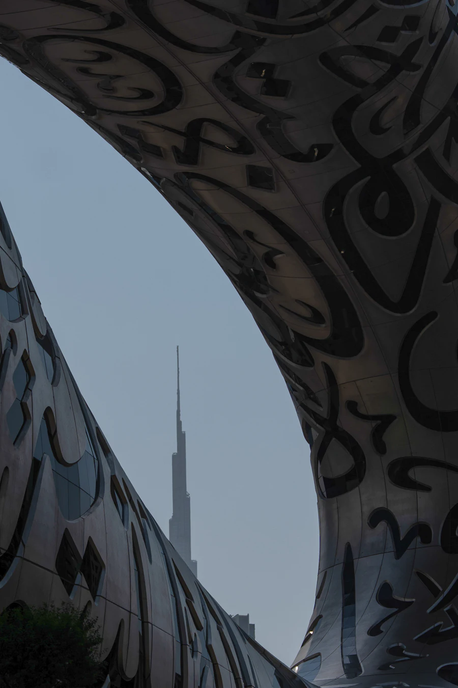 The Burj Khalifa rises in the distance, framed by sweeping, dark sculptural building facades against a pale sky. The scene emphasizes the dramatic architectural curves and vertical skyline contrast.