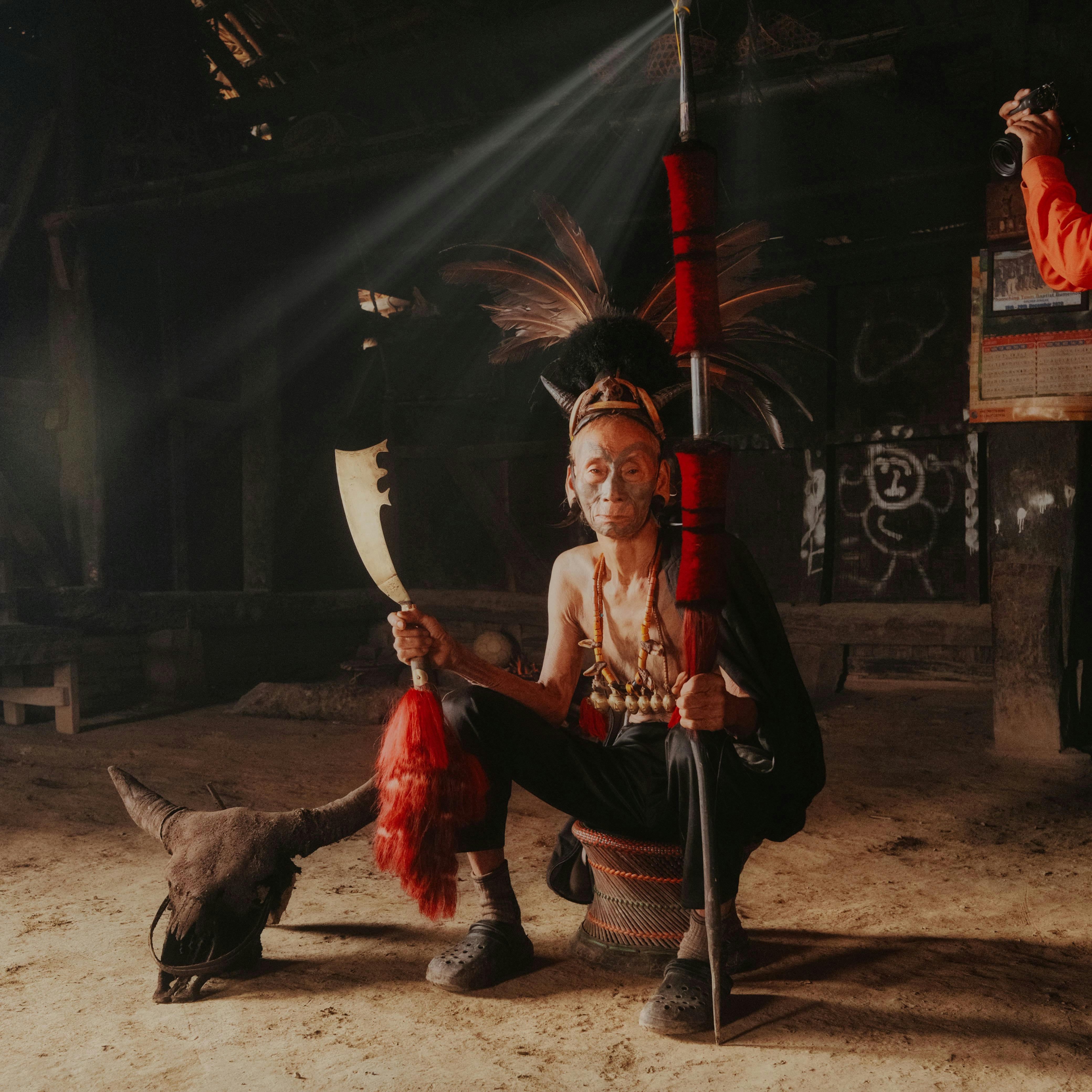 An indigenous warrior poses confidently with a traditional weapon, showcasing cultural attire and symbols in a dimly lit setting.
