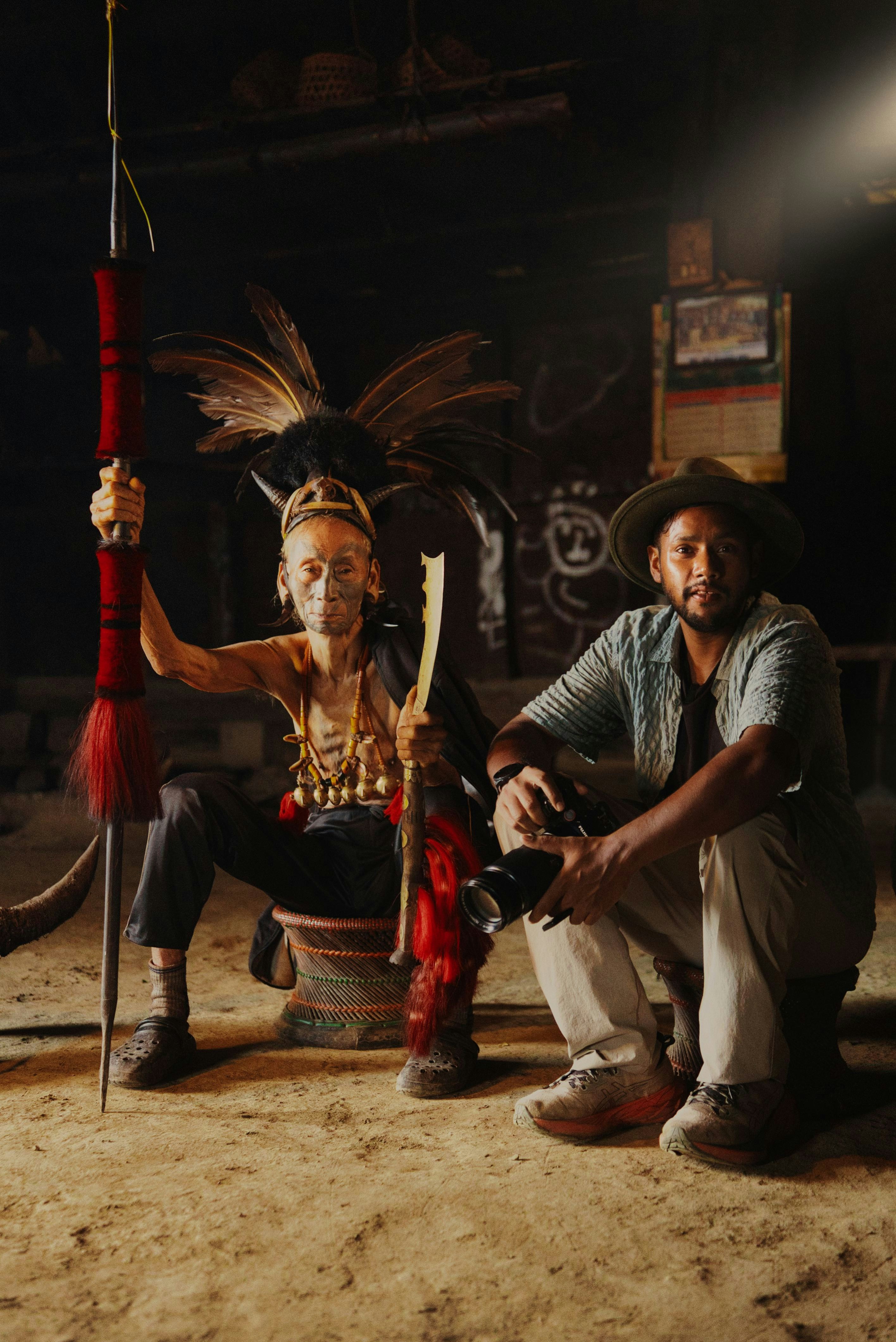 Elderly tribal leader adorned with traditional attire and artifacts poses alongside a photographer in a dimly lit environment.