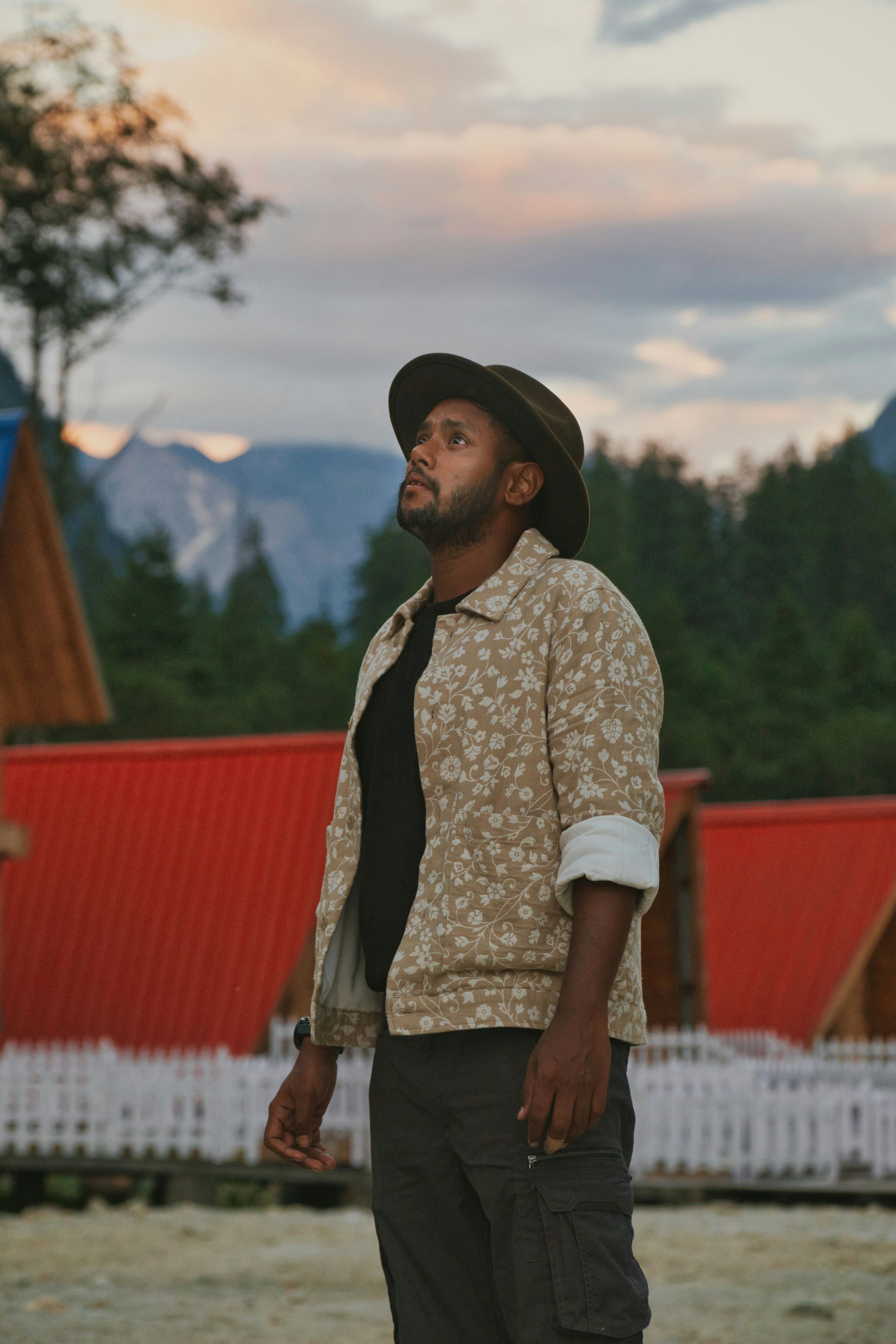 Man in a patterned shirt and hat gazes upward against a backdrop of mountains and cabins at dusk.