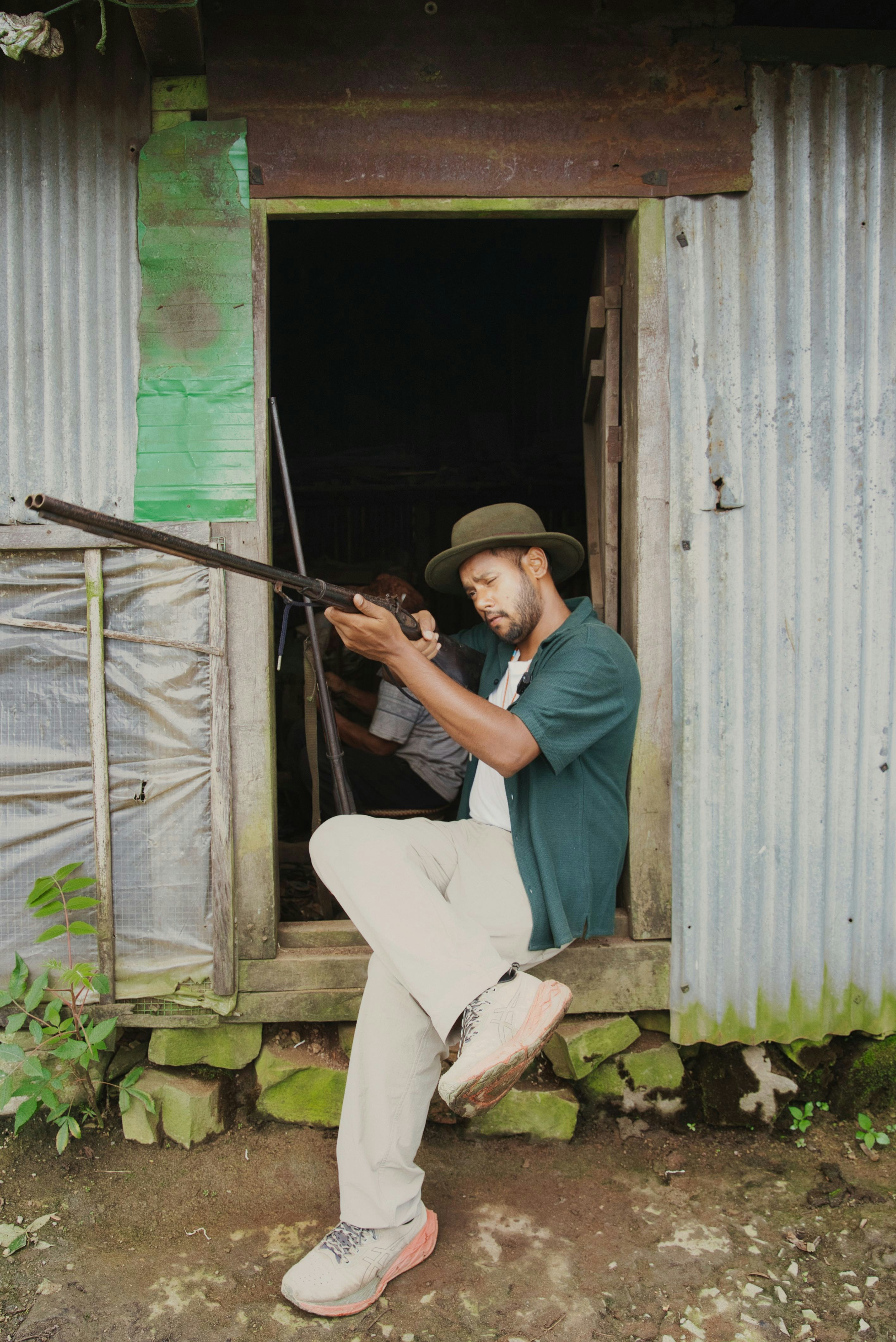 Man in hat aims rifle outside rustic building
