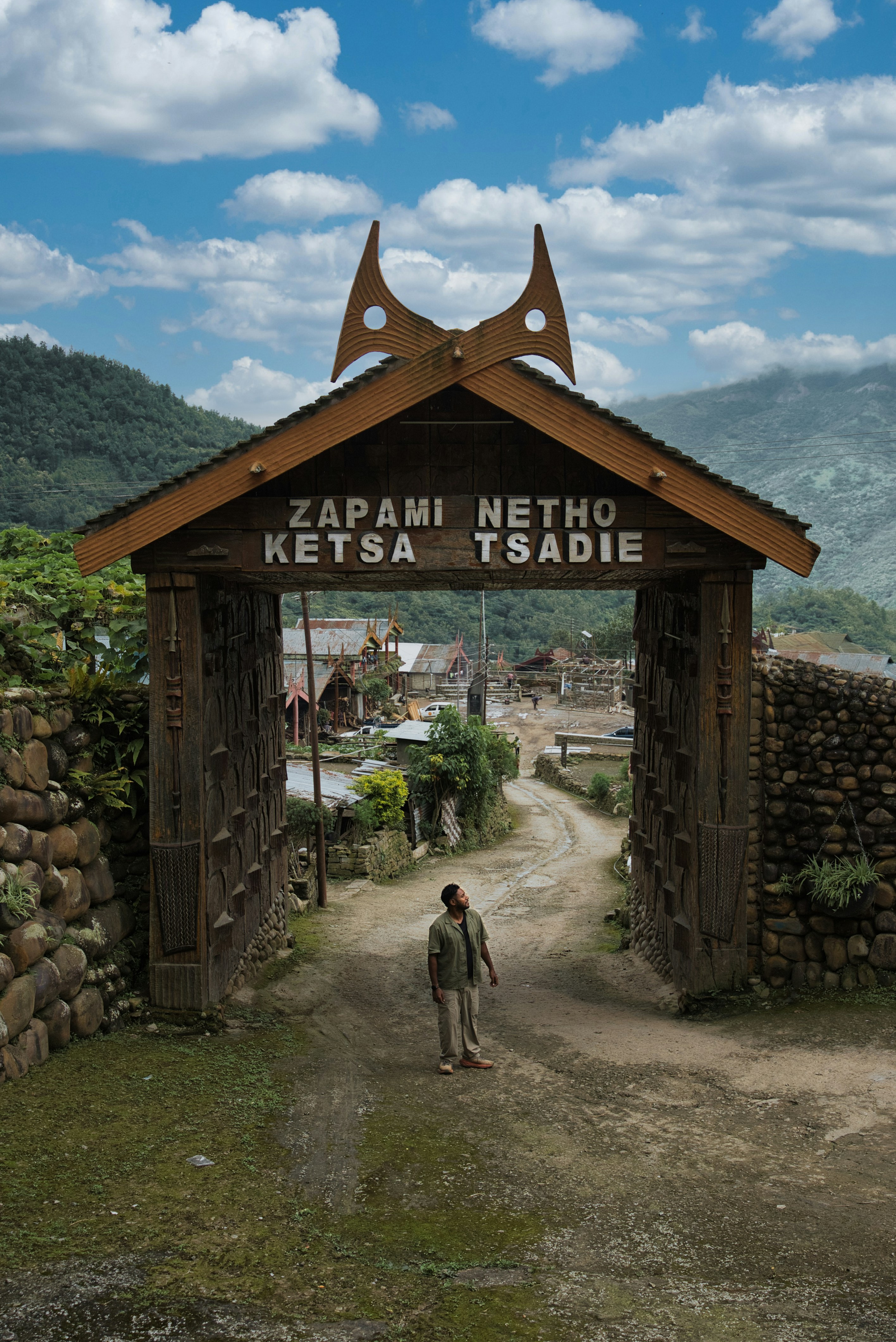 Intricately carved wooden archway welcoming visitors to a traditional village, with a solitary figure standing in contemplation on a winding path.
