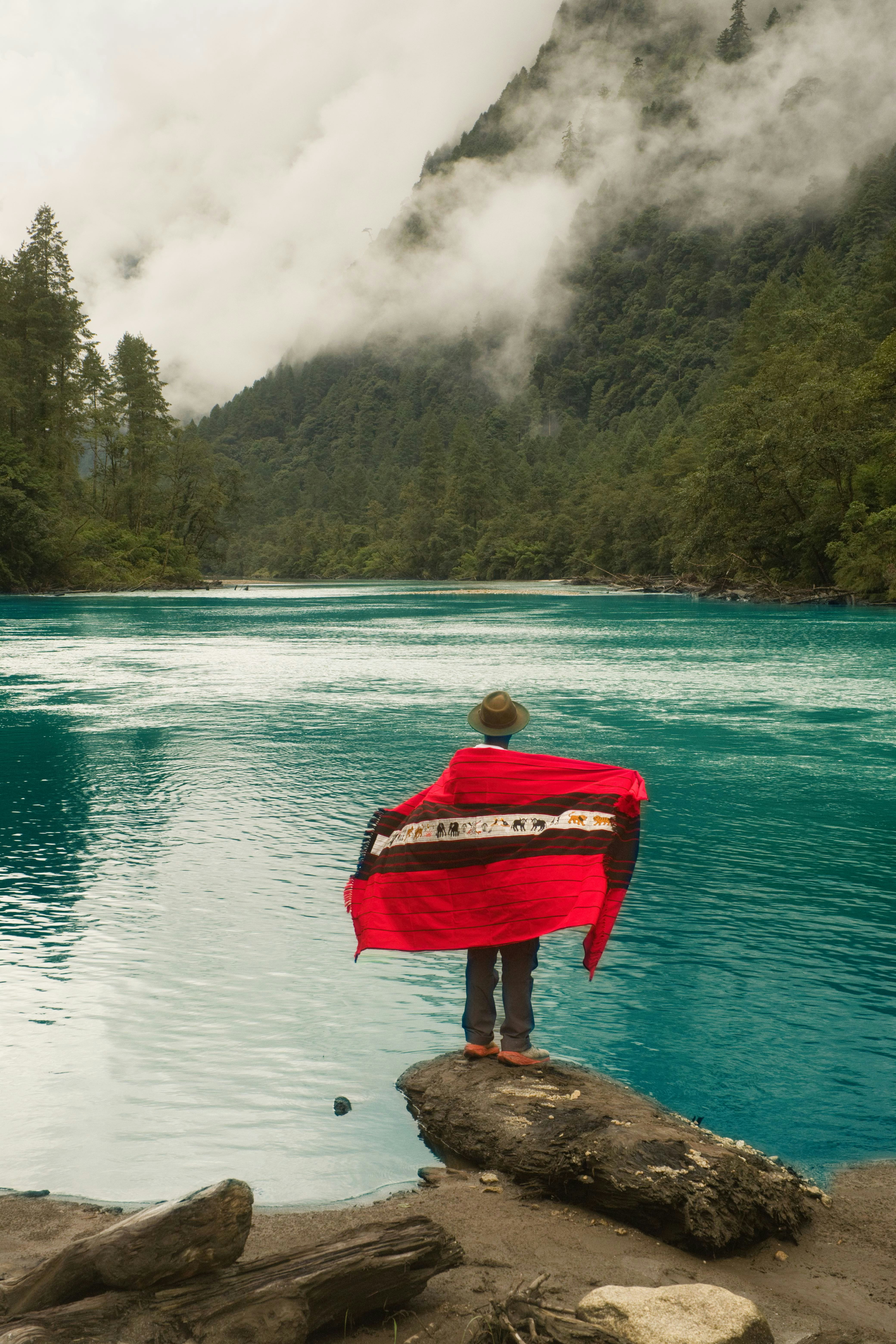 A figure draped in a vibrant red cloak stands on a log, gazing over a turquoise river framed by lush greenery and misty mountains.