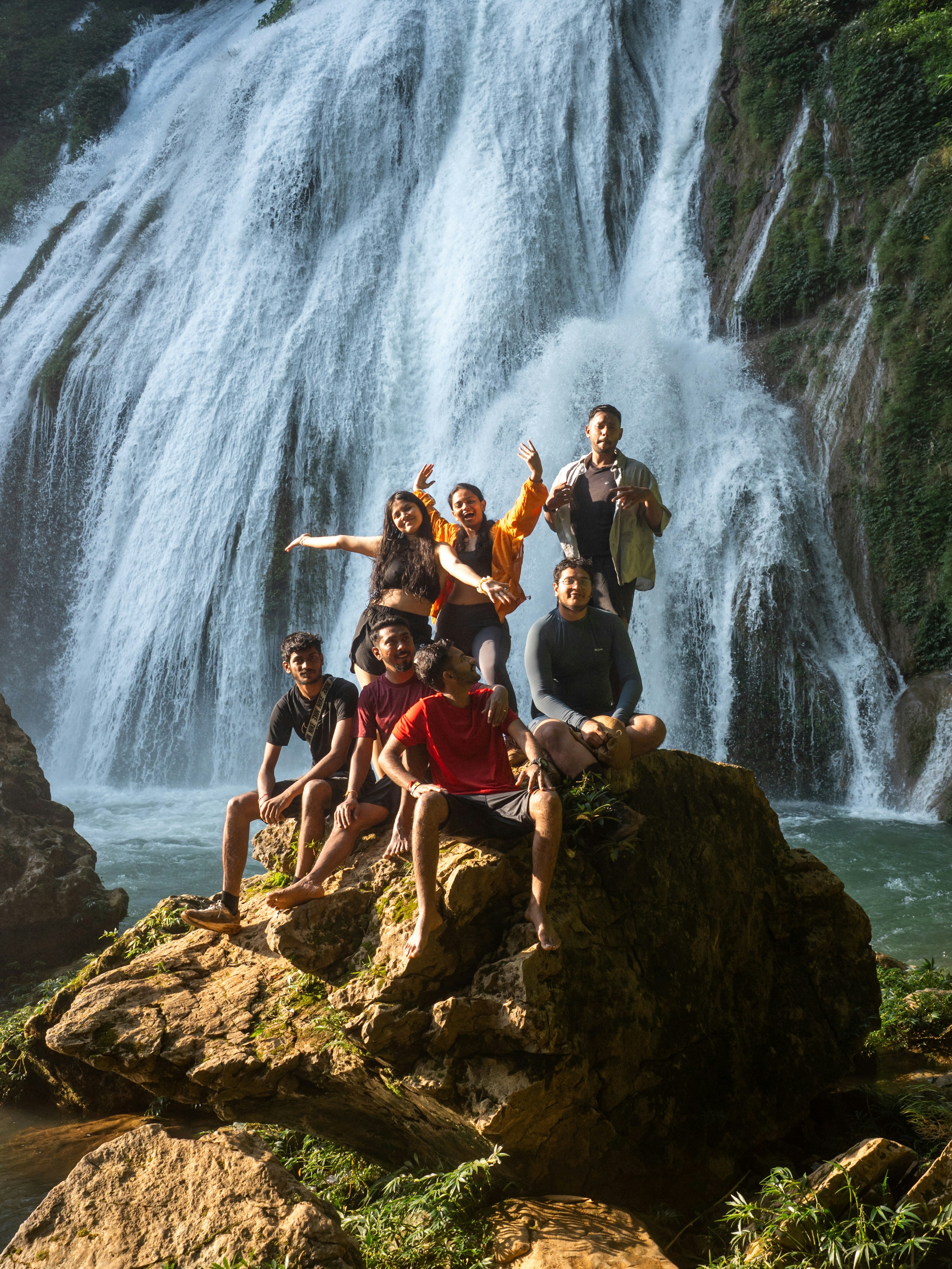Group of friends celebrating at the base of a majestic waterfall, surrounded by lush greenery and vibrant sunlight.
