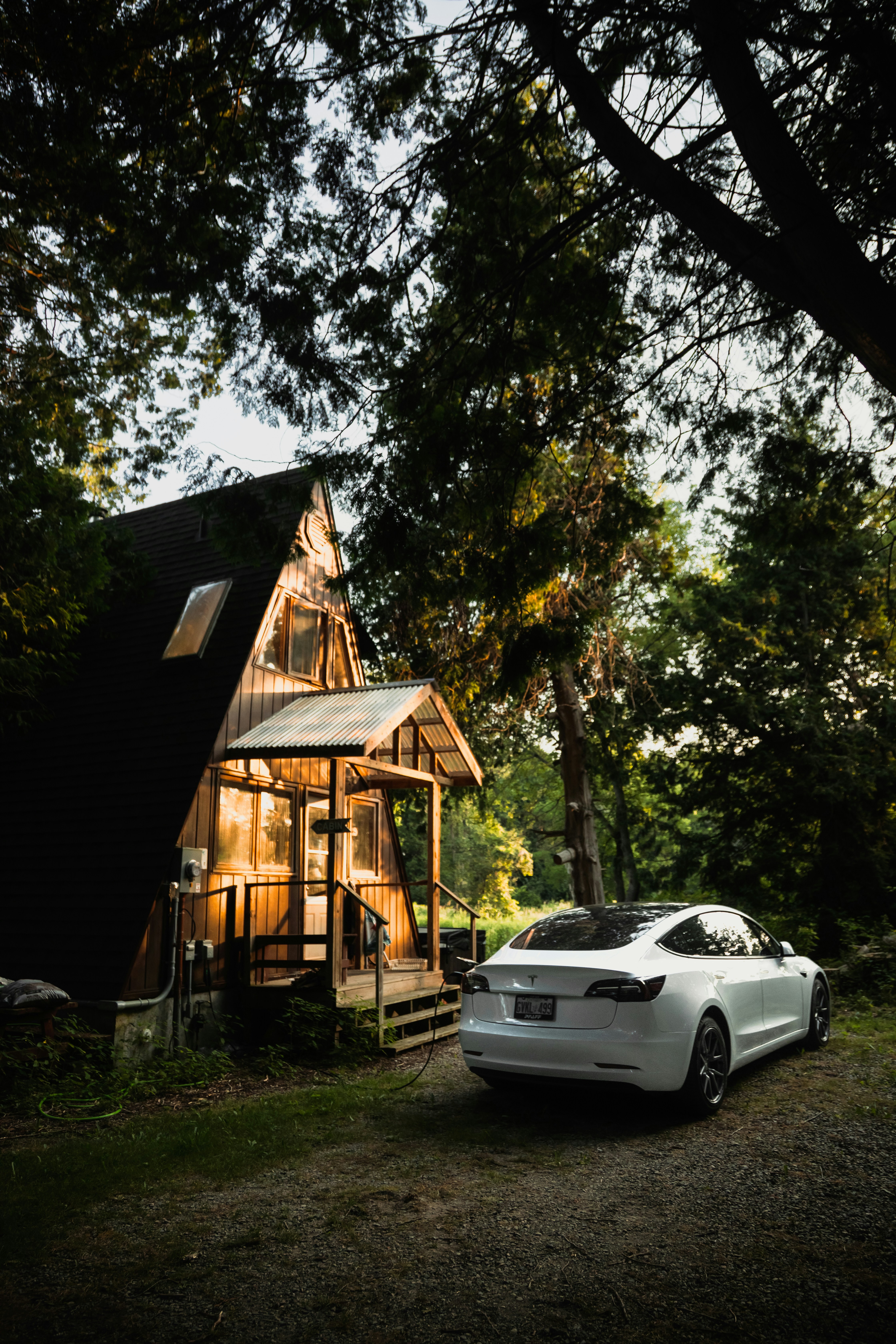 Charming wooden cabin illuminated by warm sunlight, accompanied by a sleek white electric car in a serene forest setting.
