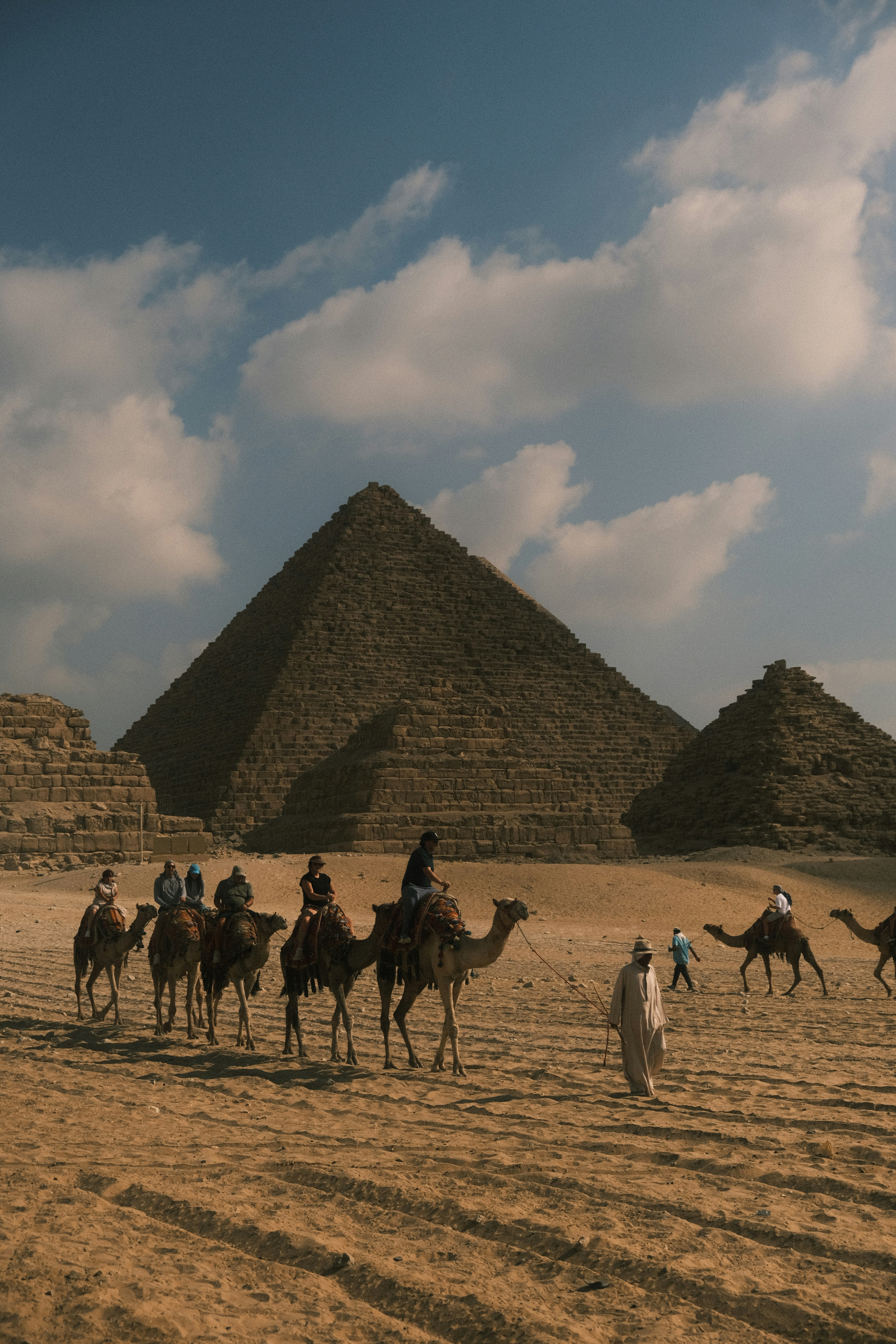 A caravan of camels traverses the sandy landscape near the majestic pyramids, with clouds softly drifting above. The scene captures the essence of ancient history and culture.