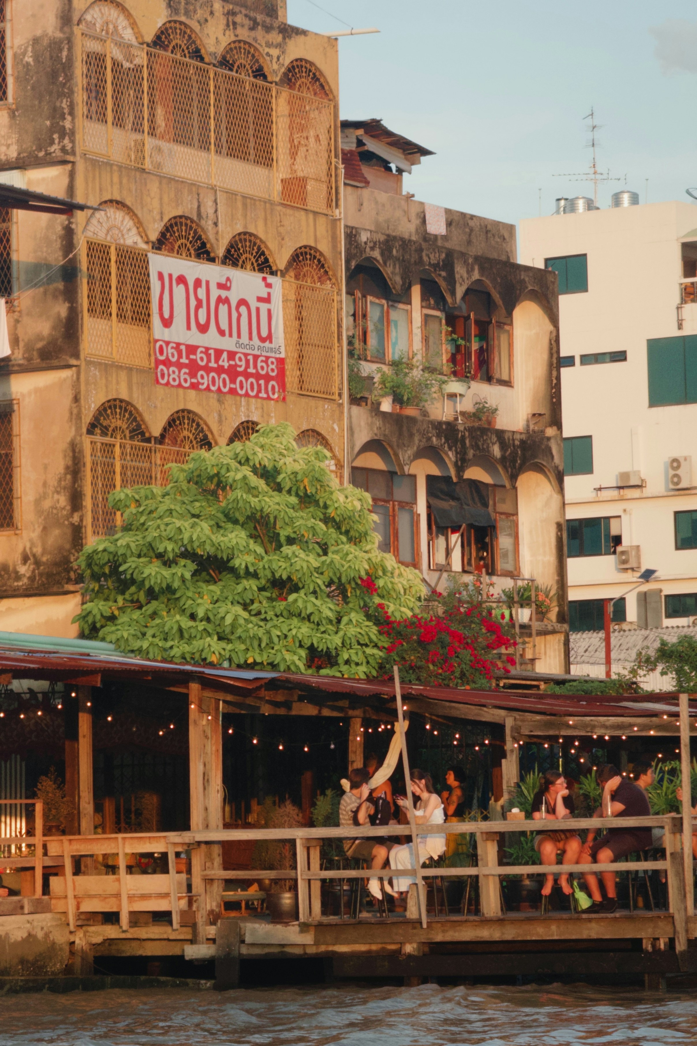 Charming riverside restaurant bustling with patrons, framed by lush greenery and vibrant flowers against a backdrop of historic architecture.