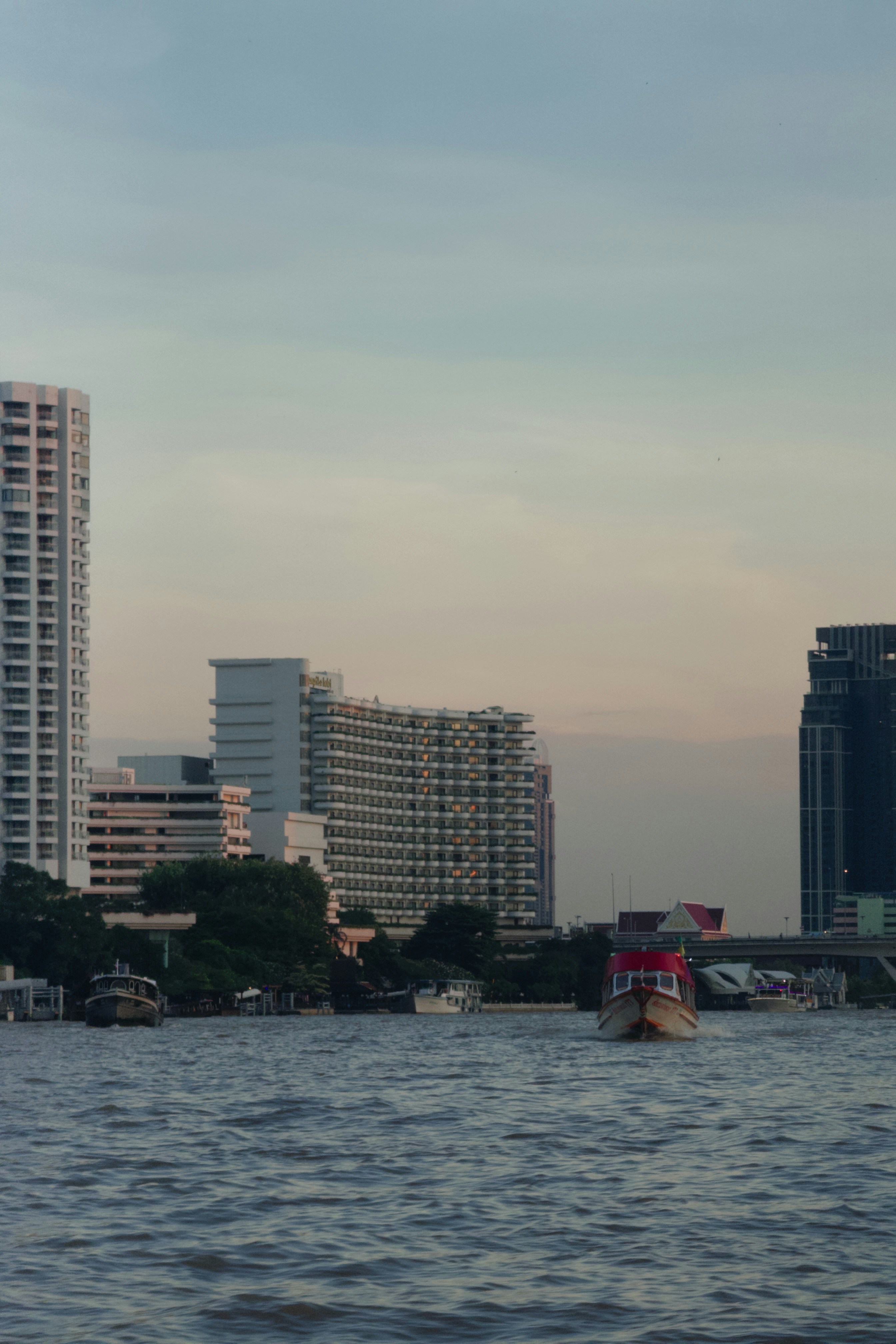 A tranquil river scene with boats gently floating in the foreground, framed by modern city buildings against a soft twilight sky.
