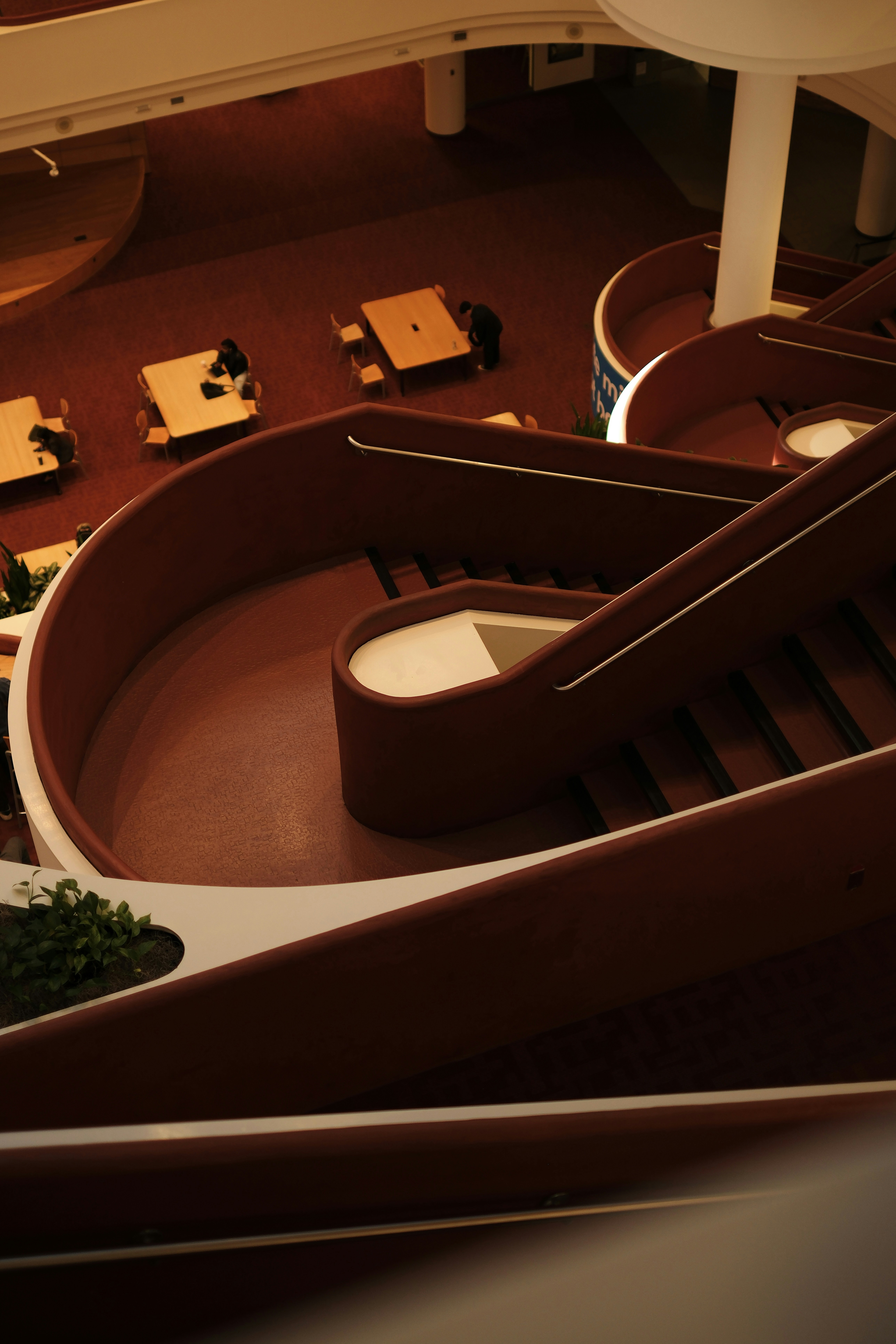 Spiral staircase within a contemporary building, showcasing smooth lines and warm tones. Tables and people are visible in the background, adding context to the space.