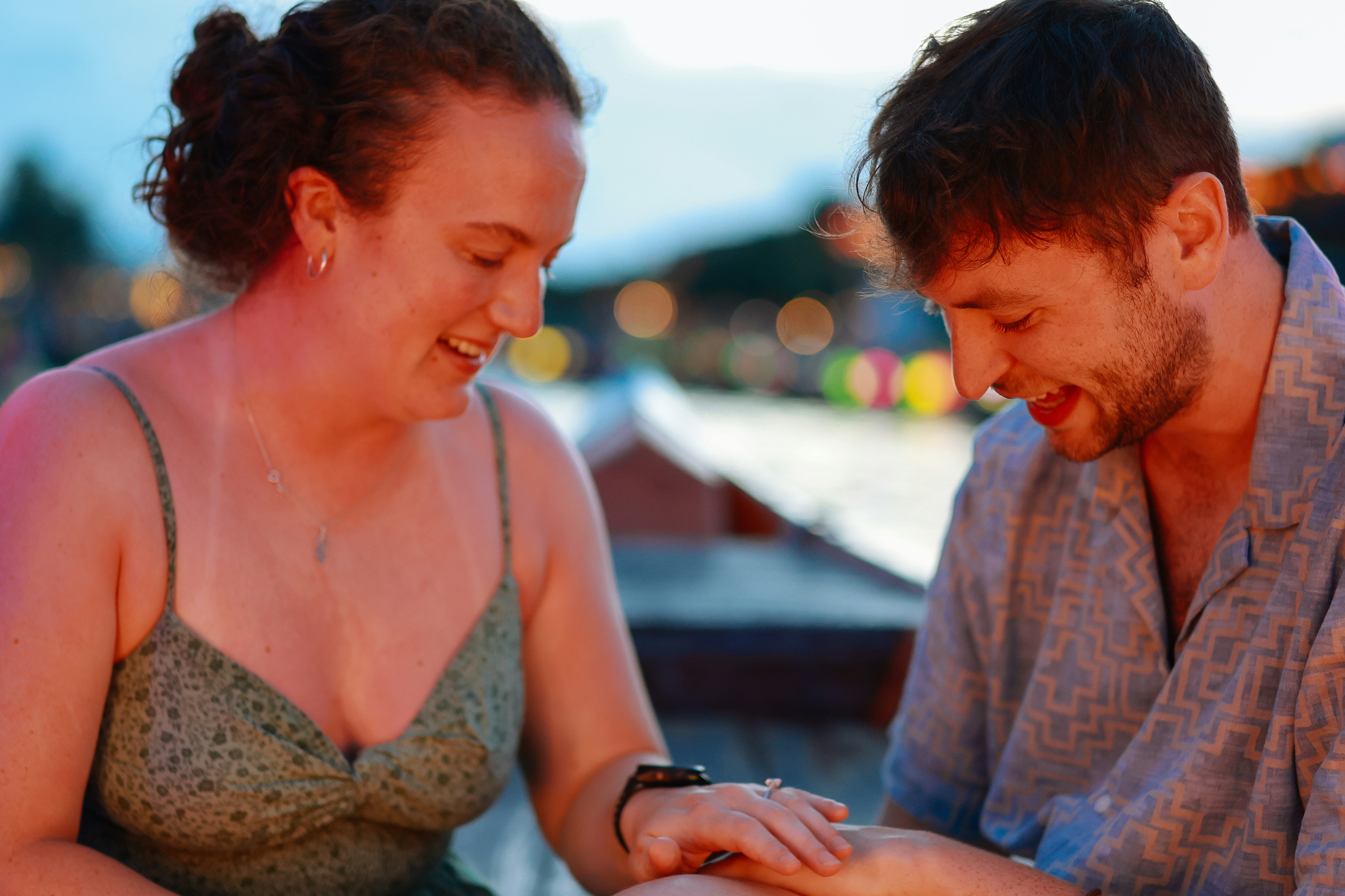 Couple sharing a joyful moment on a waterfront, with soft bokeh lights in the background. The scene captures their intimate interaction beautifully.