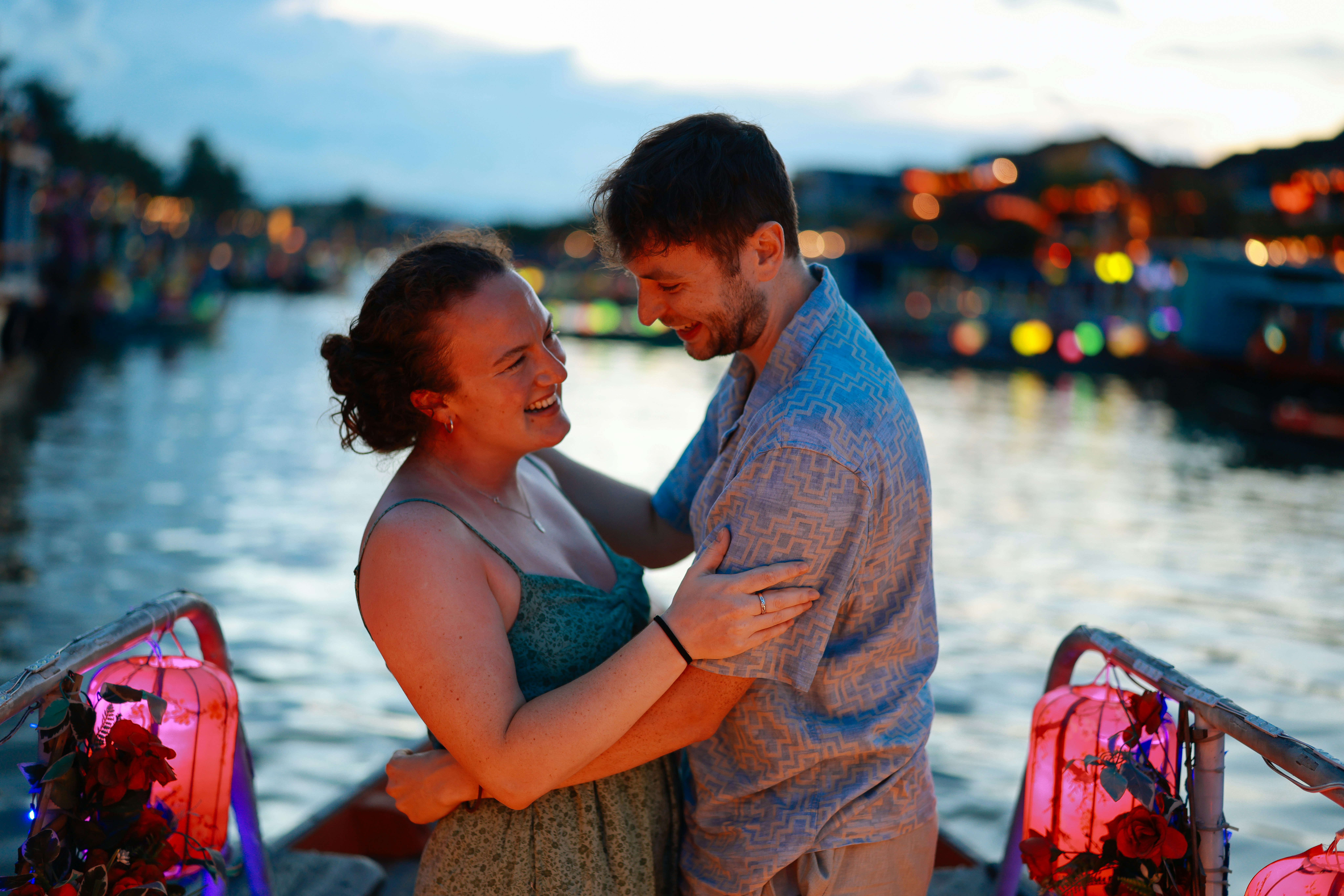 Couple joyfully sharing a moment on a boat, surrounded by colorful lights reflecting on the water. The scene captures the warmth of their connection against a serene twilight backdrop.