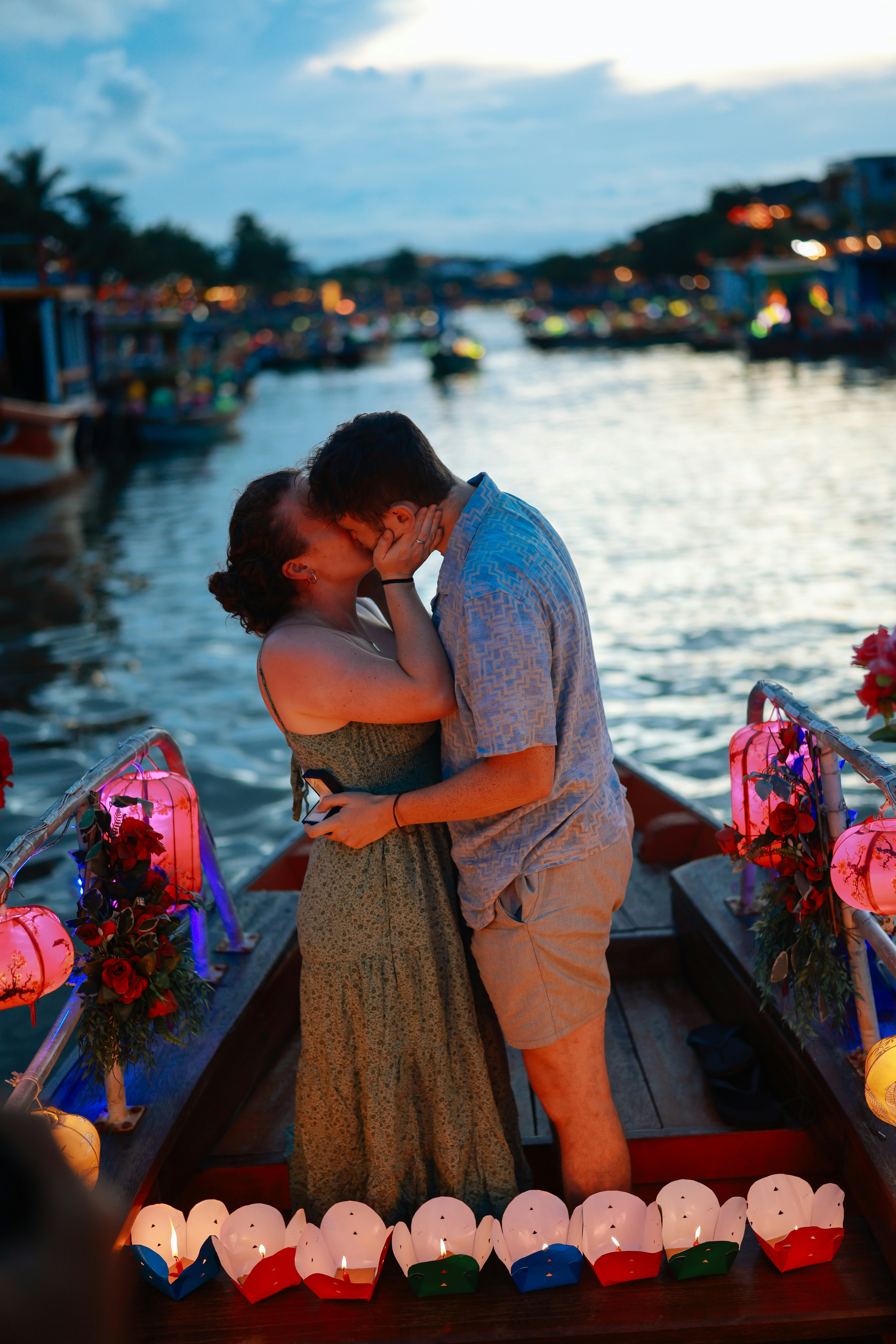 Couple sharing a kiss on a boat adorned with colorful lanterns, surrounded by a shimmering river at twilight.