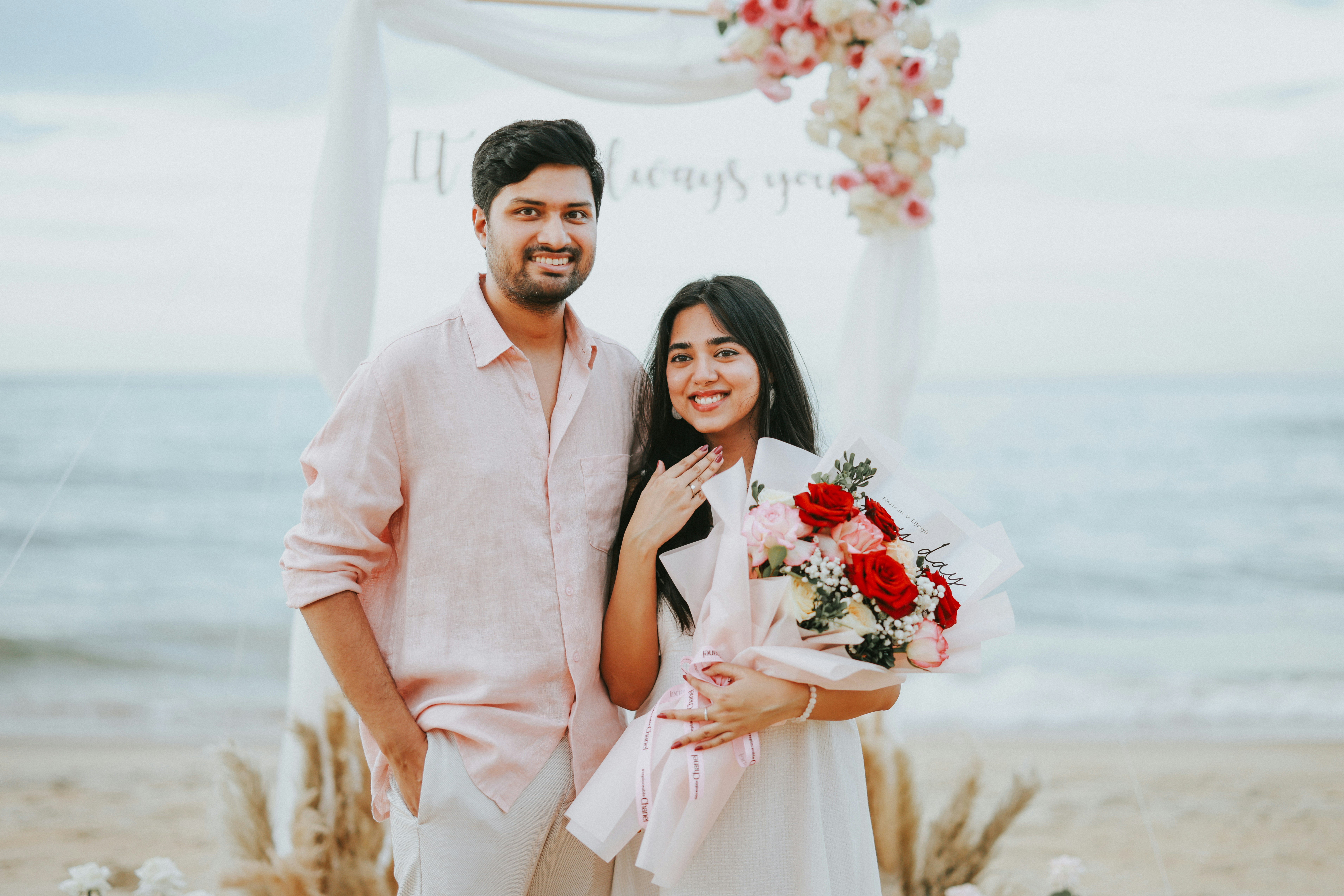 Couple celebrating a special occasion on the beach, surrounded by floral decorations and holding a bouquet of roses.