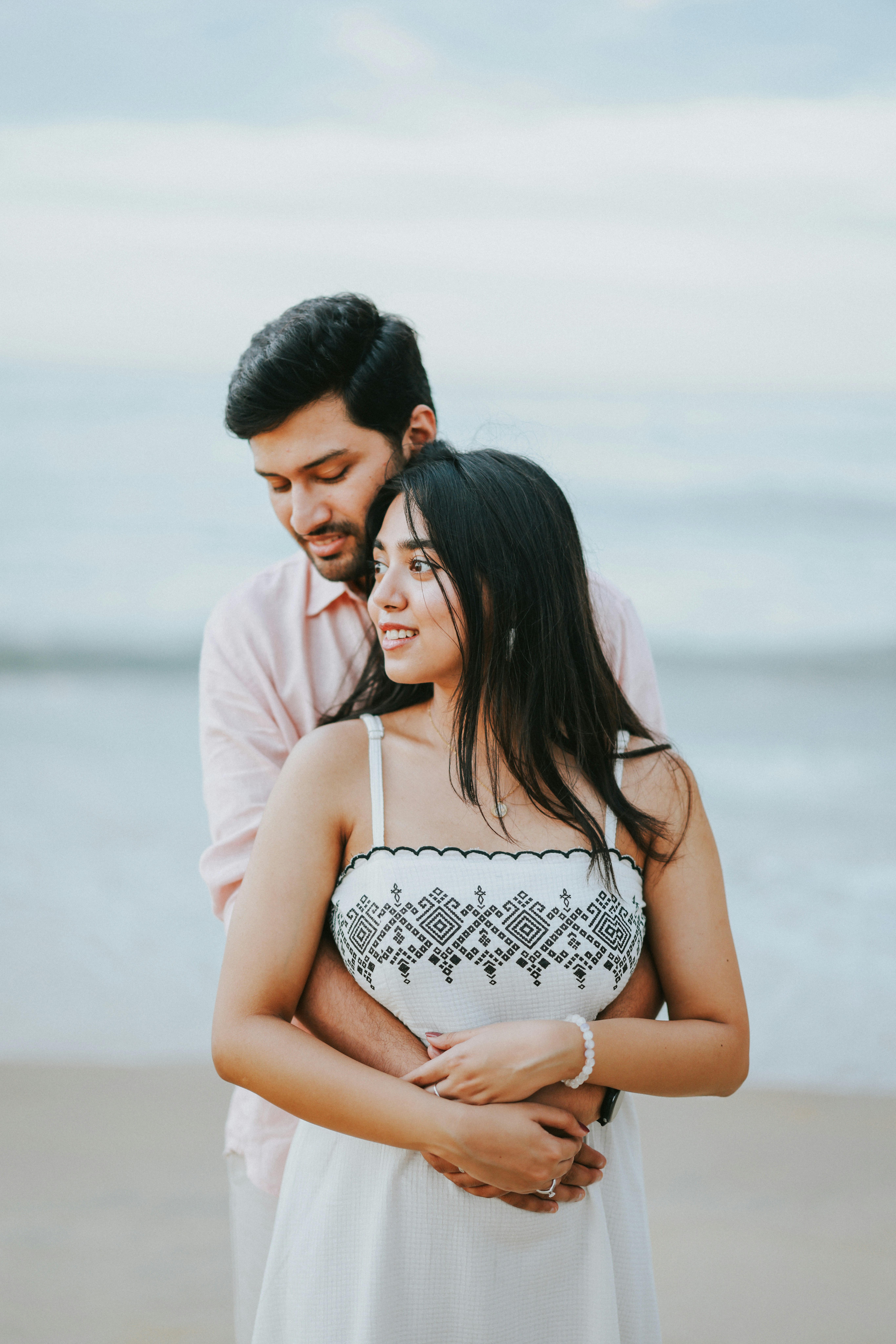 Couple sharing a tender moment on the beach, with gentle waves in the background. The woman wears a white dress adorned with intricate patterns.