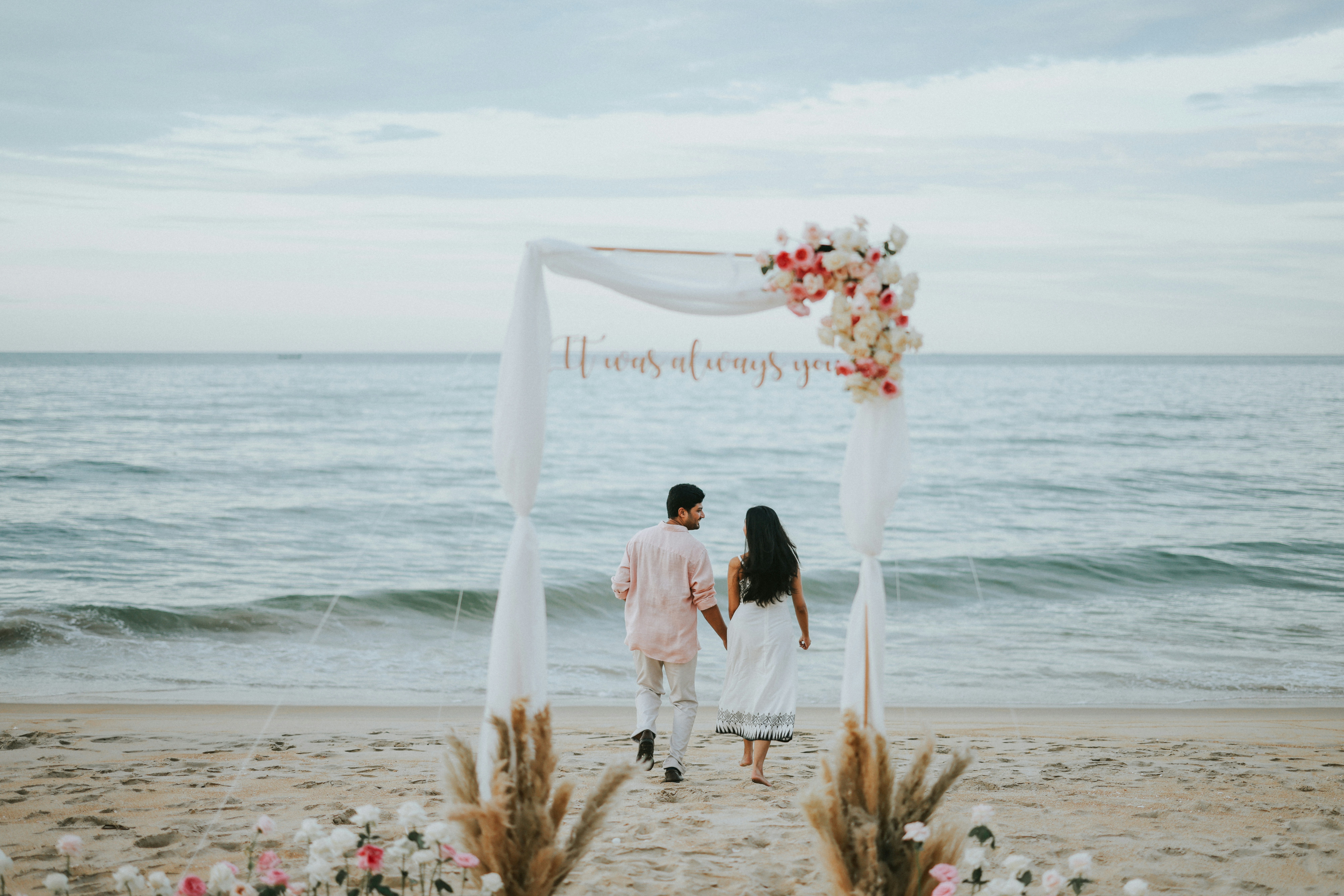 Couple walking hand-in-hand under an arch adorned with flowers, heading towards the tranquil sea. The scene captures a romantic moment at the beach.
