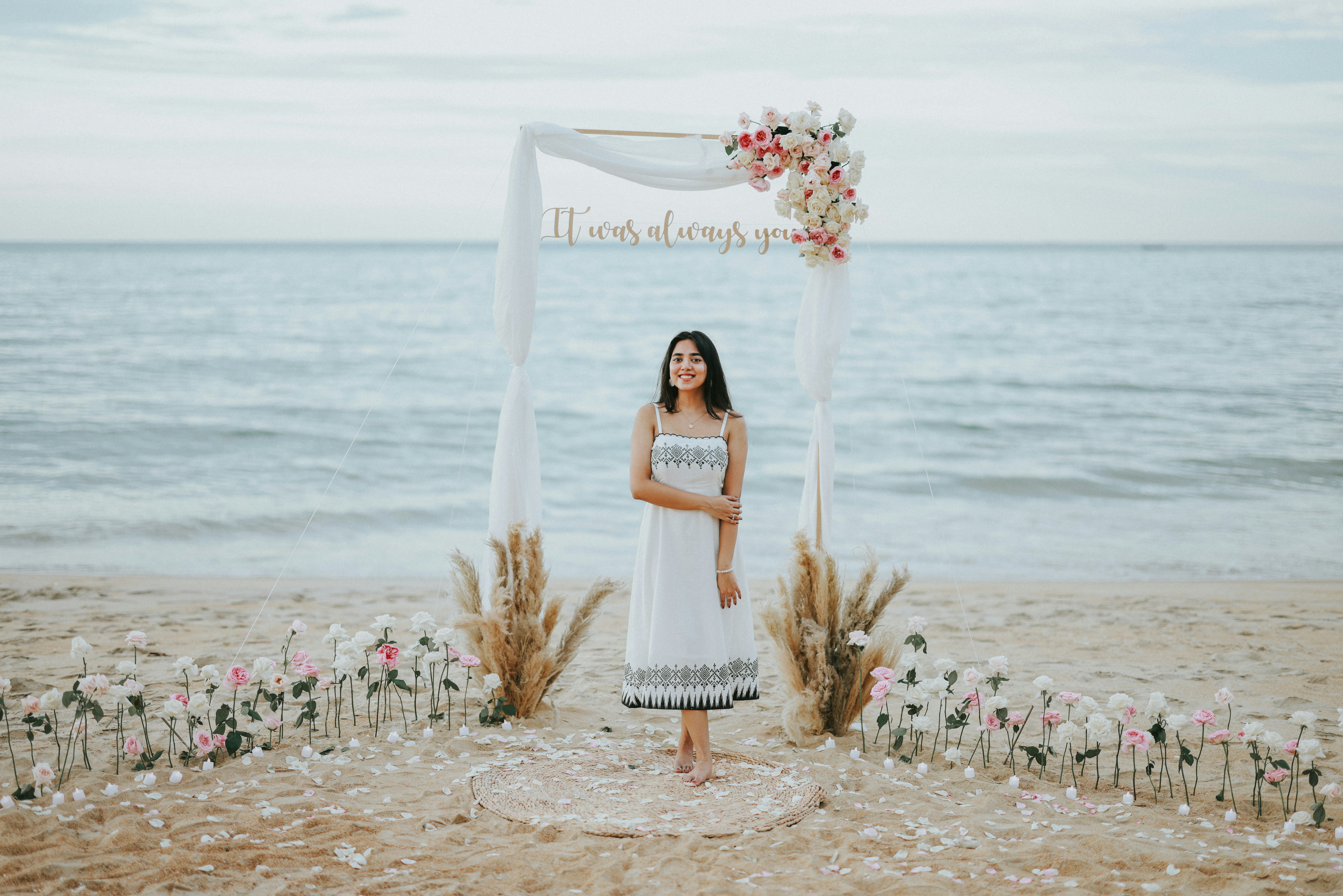 A woman in a white dress stands gracefully beneath a floral arch on a sandy beach, with gentle waves in the background. The scene is adorned with petals and greenery, creating a romantic atmosphere.