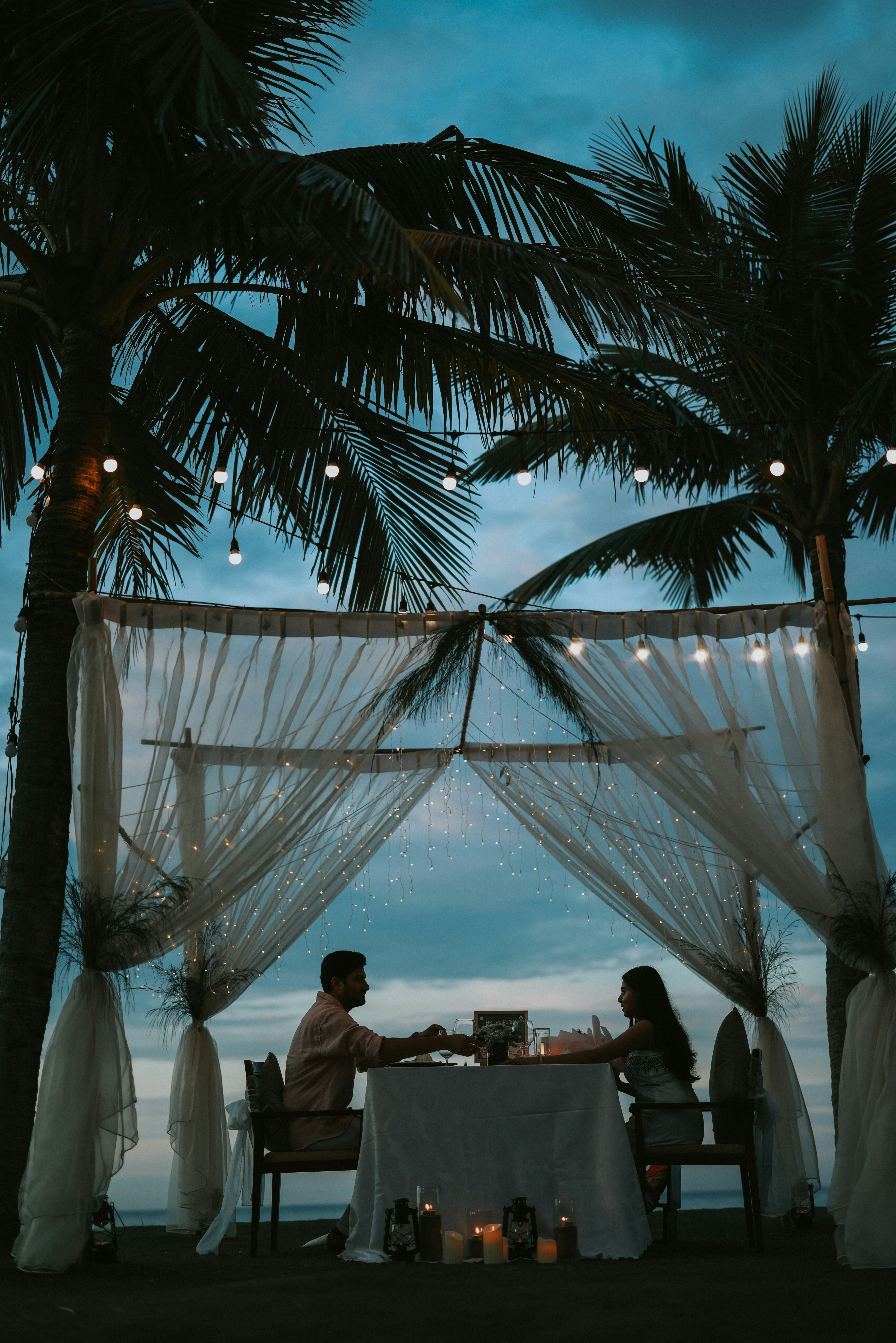 Couple sharing a romantic dinner beneath a canopy of lights and palm trees at twilight.
