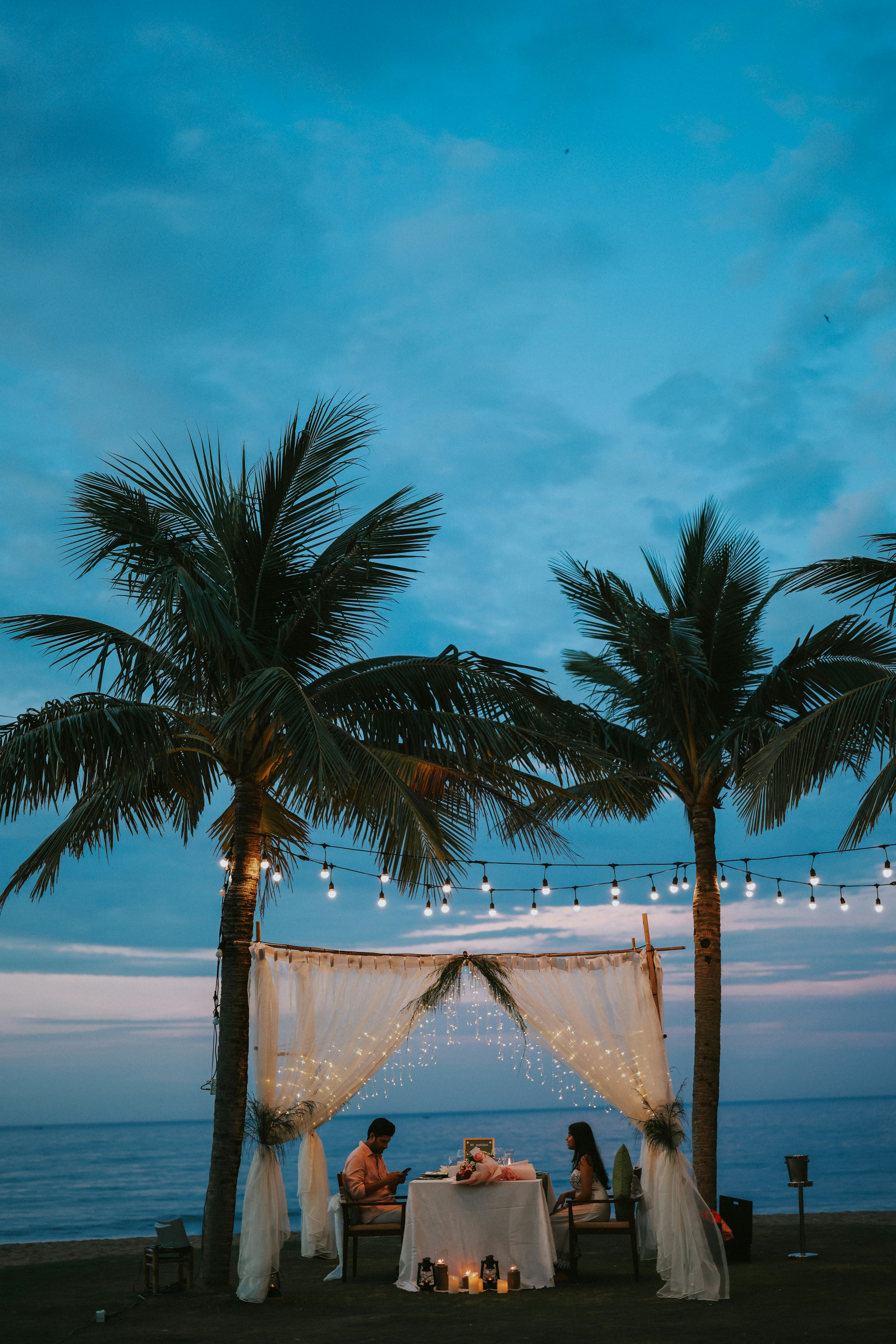 Couple enjoying a romantic dinner setup on the beach, surrounded by palm trees and fairy lights. The ocean waves gently lap at the shore in the background.