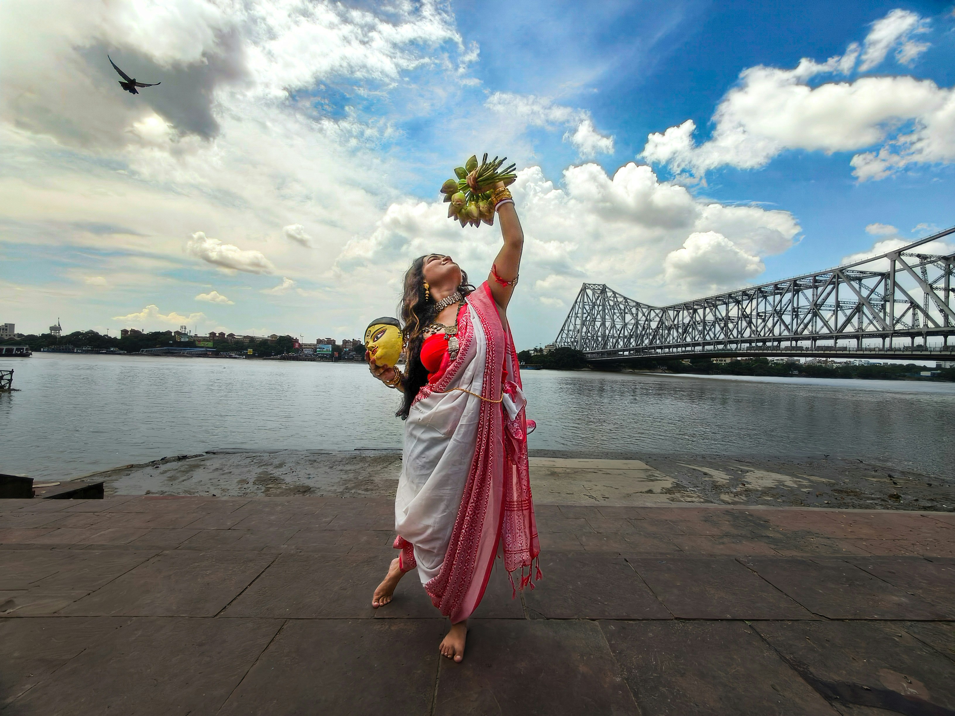 Woman in traditional attire gracefully poses with fruits against a river backdrop and an iconic bridge. Clouds add drama to the scene.