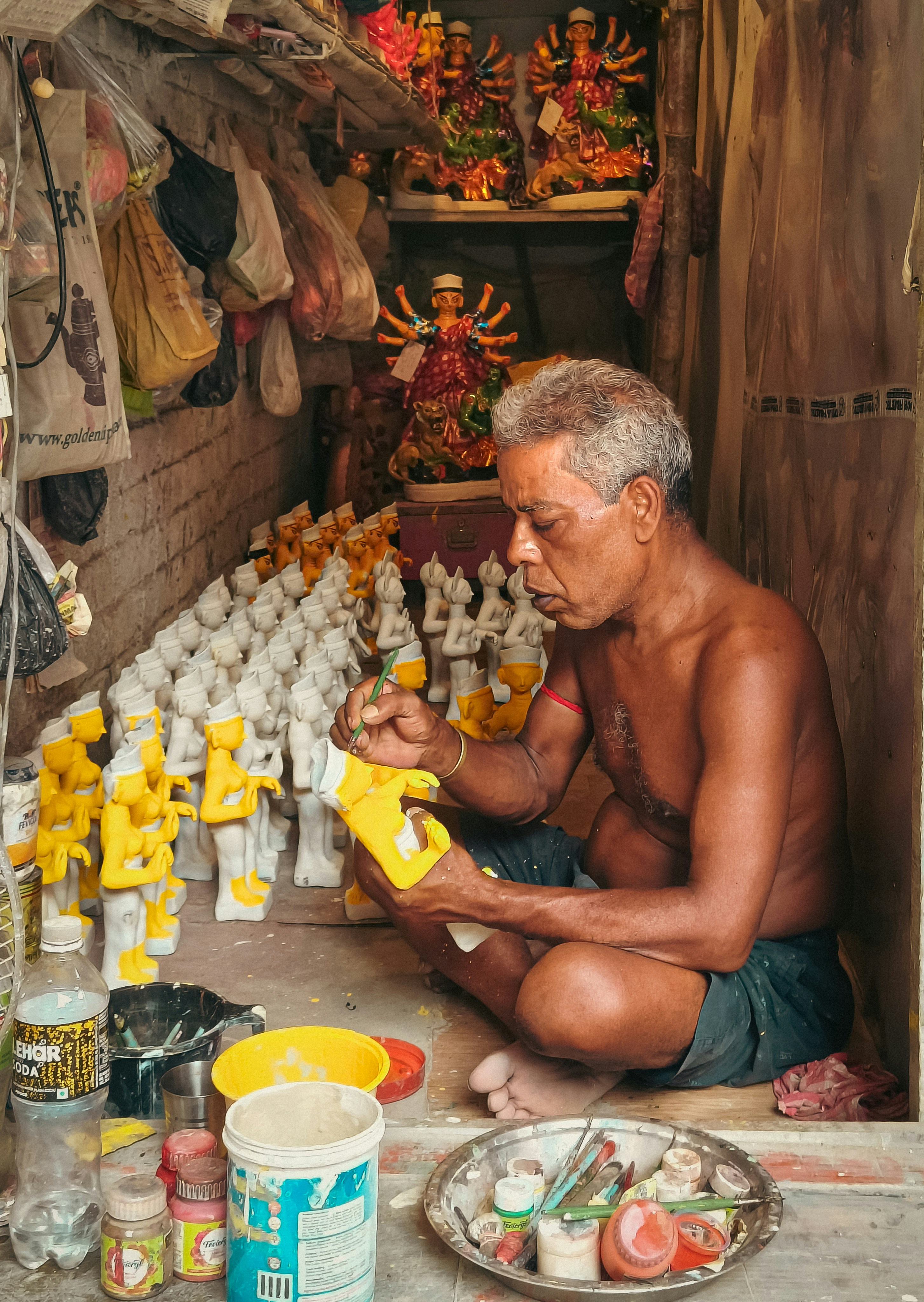 Artisan meticulously painting a yellow idol in a cramped workshop filled with partially finished figures. Colorful decorations and tools surround him.