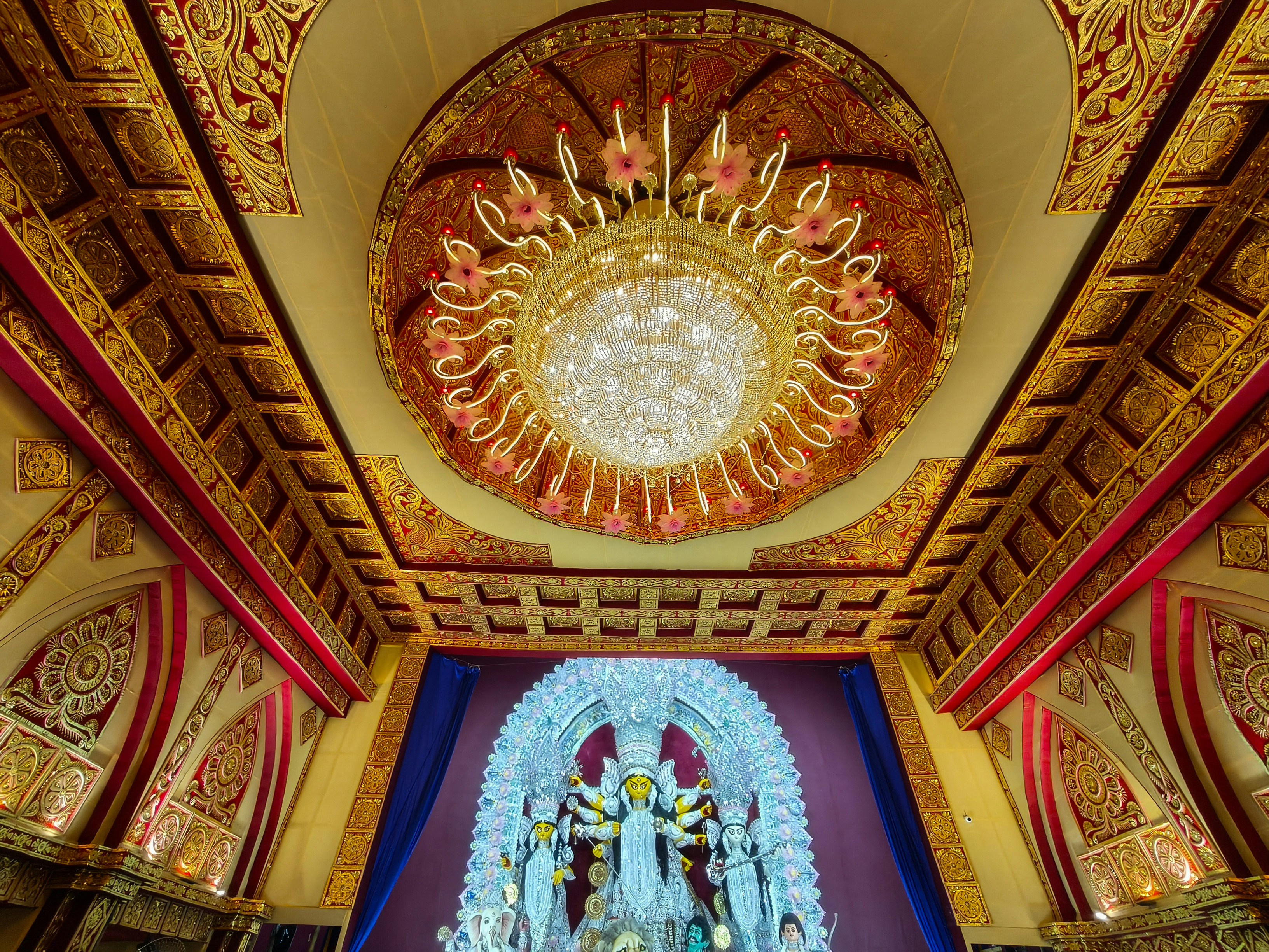 Intricate ceiling adorned with ornate patterns and a grand chandelier above a vibrant statue of a deity in a richly decorated hall.