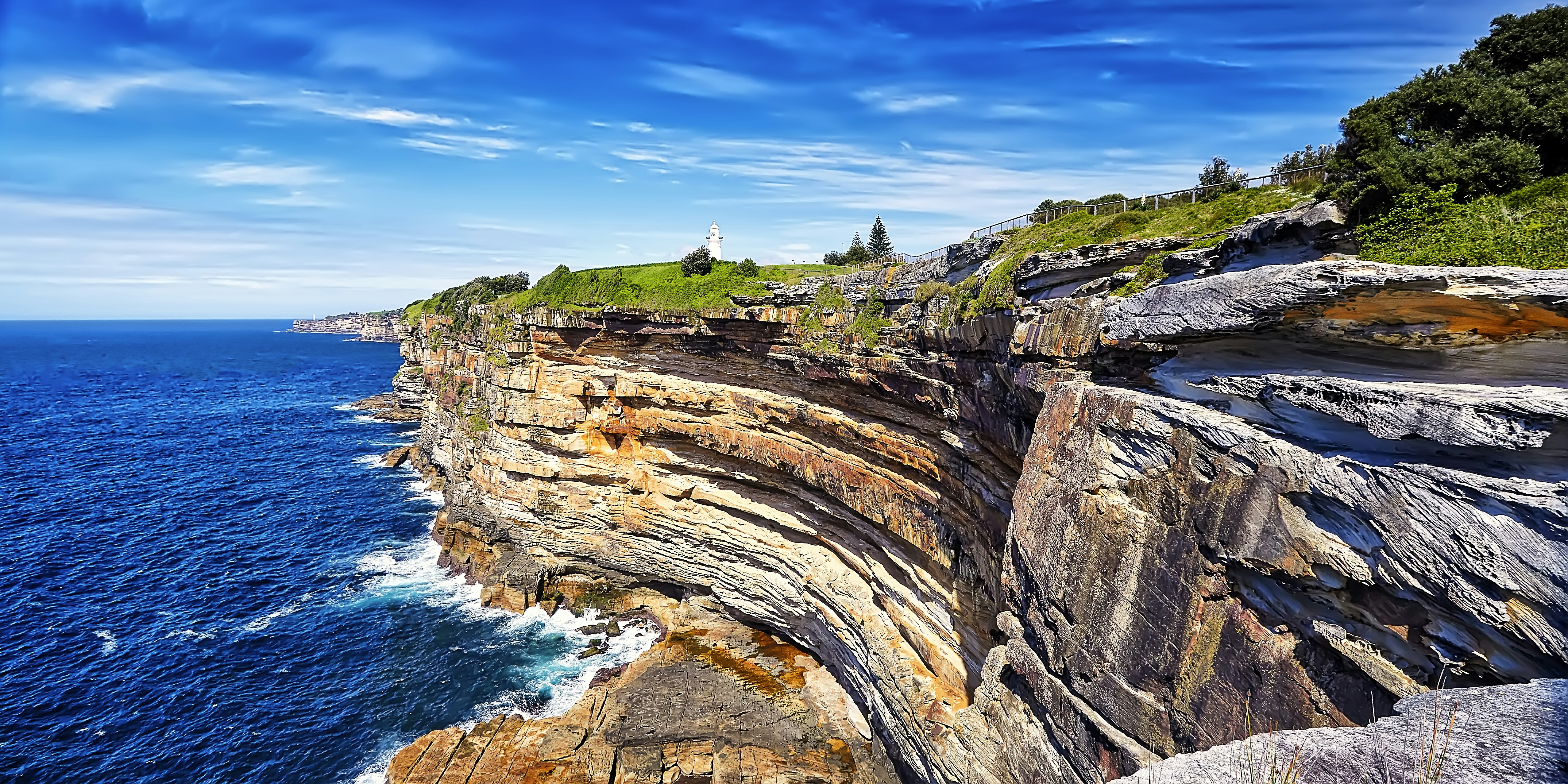 Dramatic coastal cliffs rise sharply above the deep blue ocean, with a lighthouse perched on the green hillside. The scene captures the powerful interaction between land and sea.