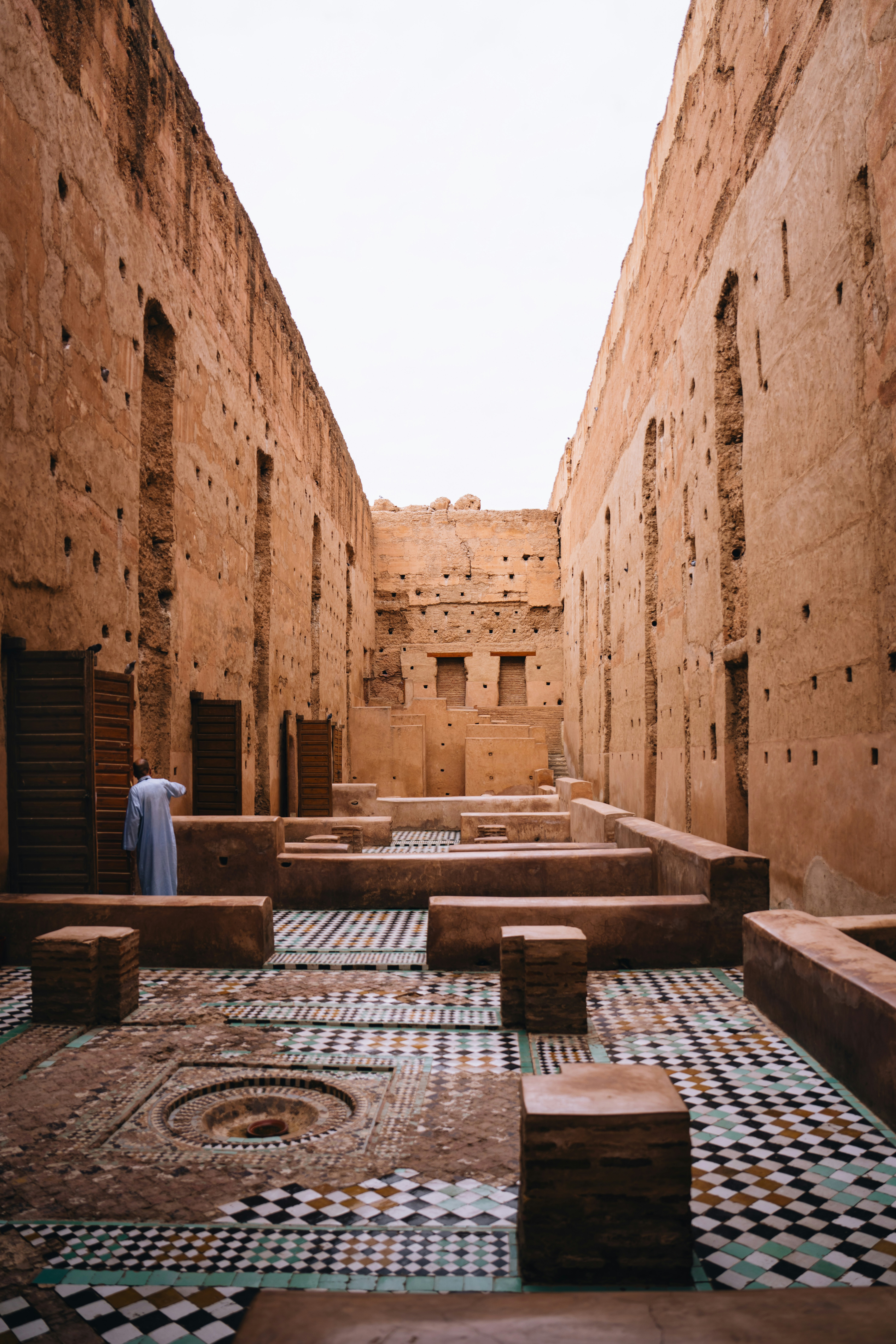A vast, ancient courtyard with textured stone walls and intricate patterned tiles, evoking a sense of timelessness. A figure in traditional attire explores the space.