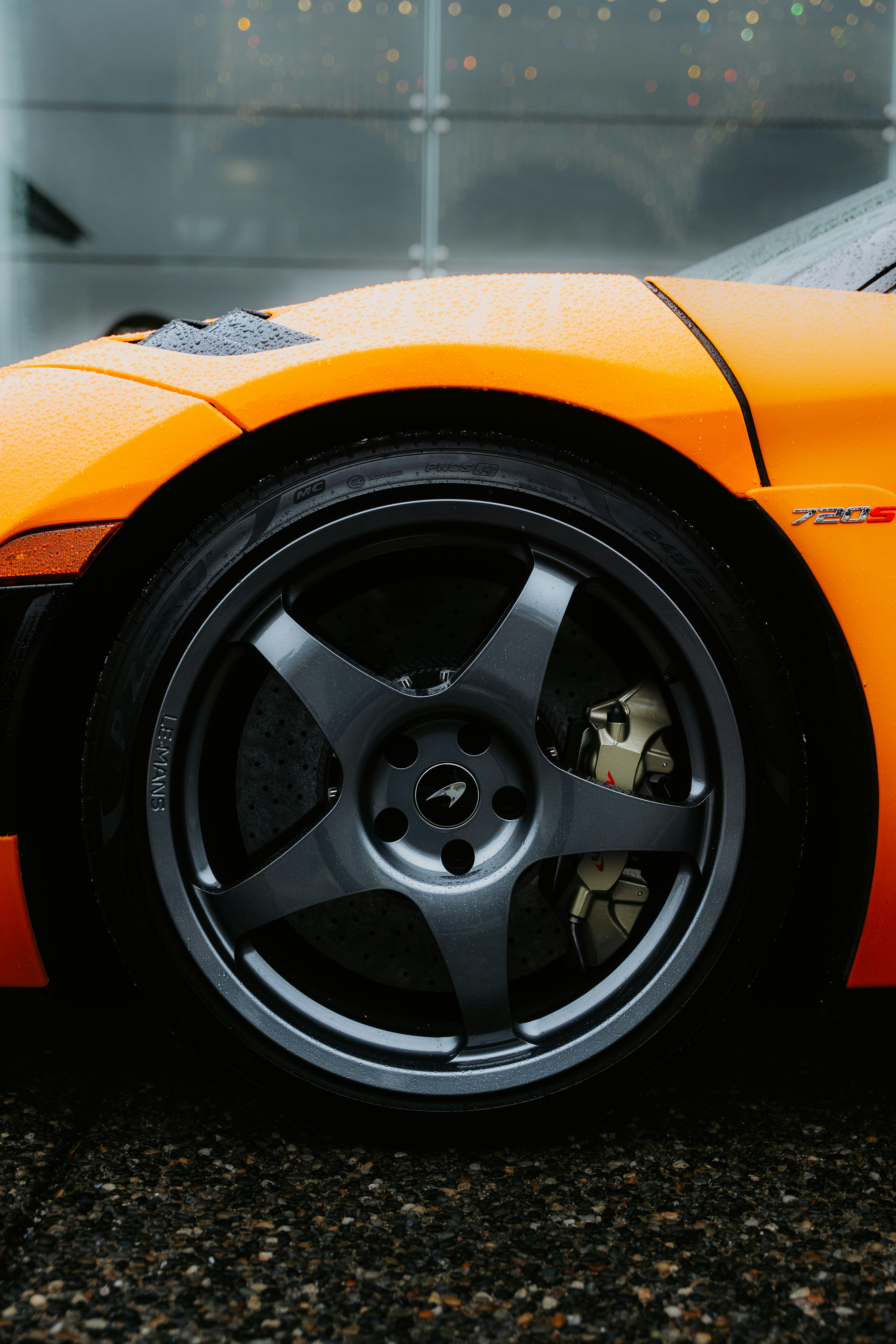 Close-up of a sleek orange sports car wheel showcasing intricate design and engineering. The wet surface adds a dynamic texture to the scene.