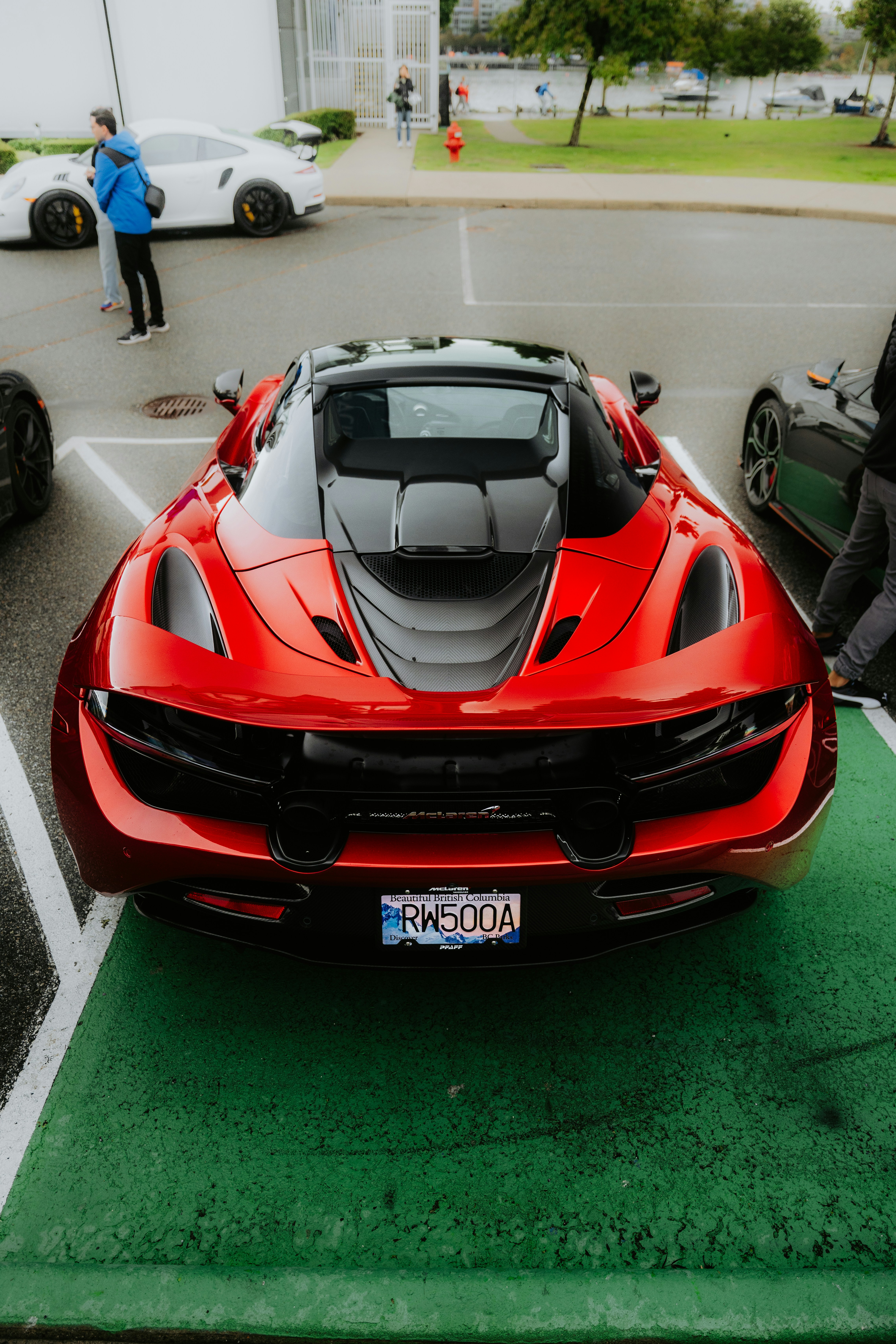 Red McLaren supercar parked on green pavement with blurred background of other vehicles and people.