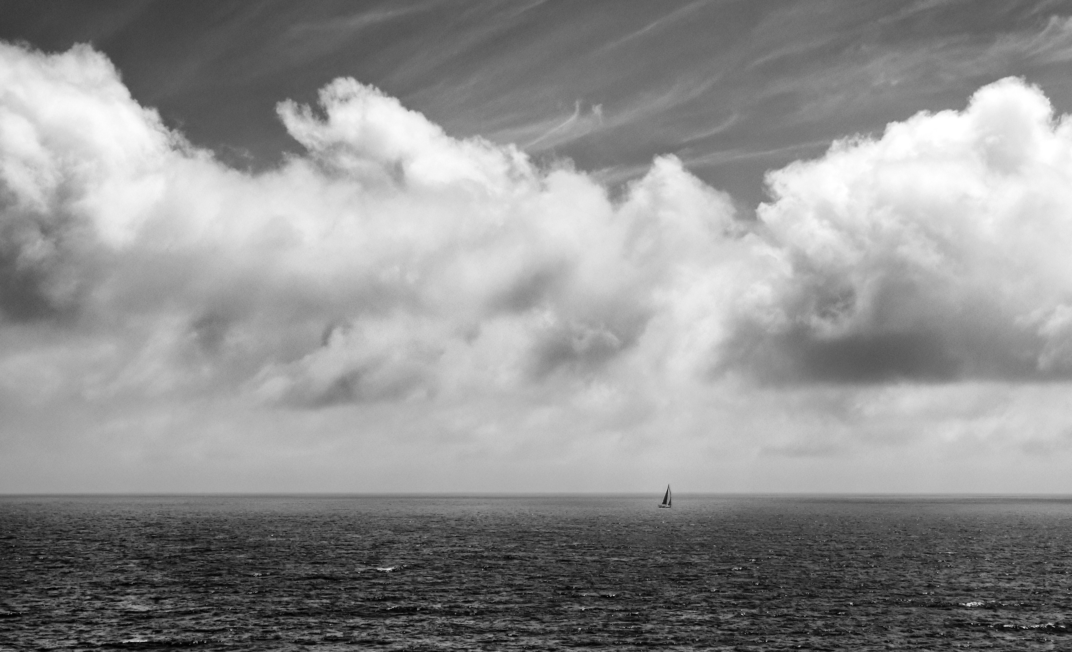 Black and white seascape with a solitary sailboat on the horizon beneath vast, textured clouds — an image of balance between movement and stillness. | A lone sailboat on the vast ocean under dramatic clouds