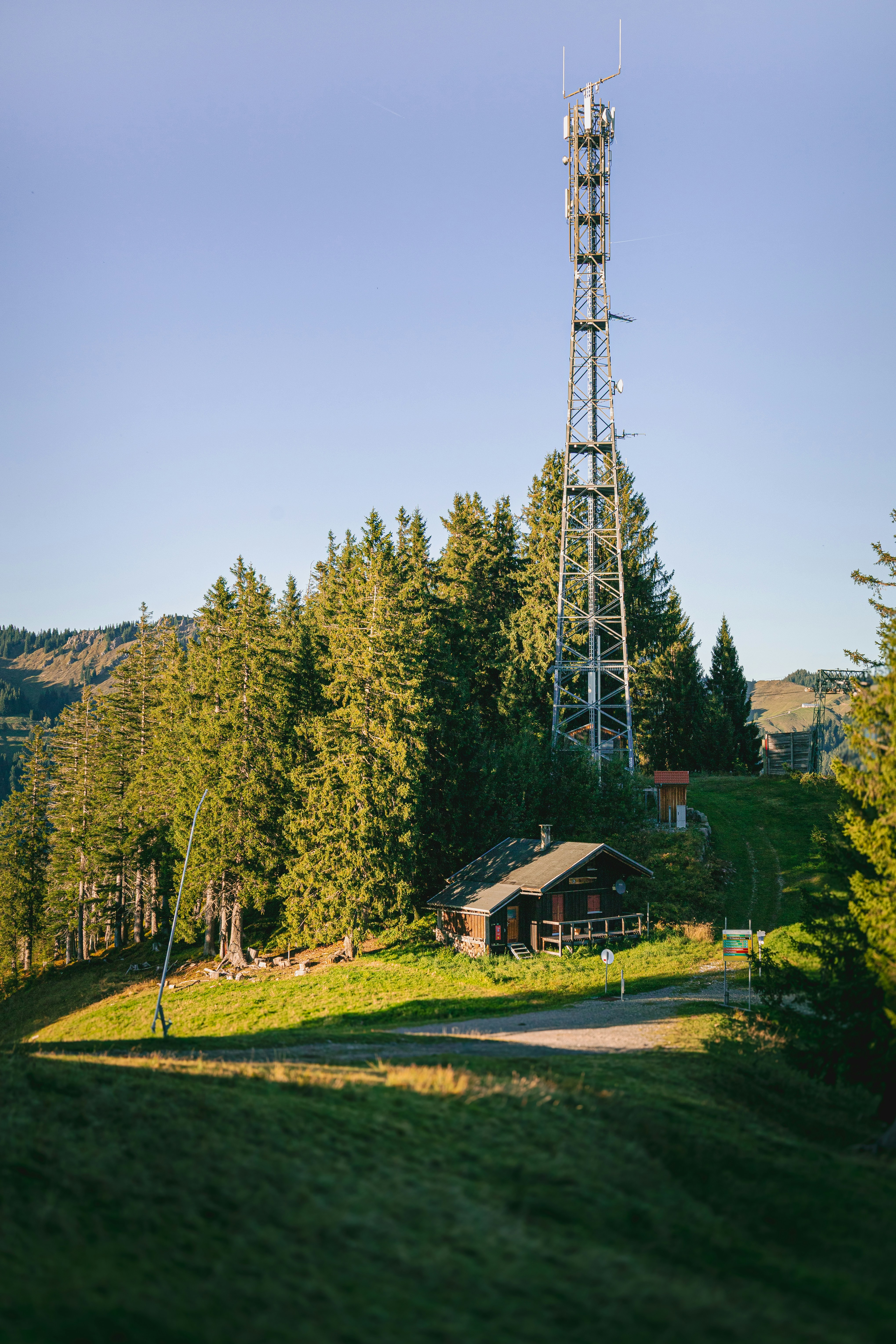 A telecommunications tower rises above a rustic cabin, surrounded by lush trees on a hillside. The scene captures the balance between nature and modern infrastructure.