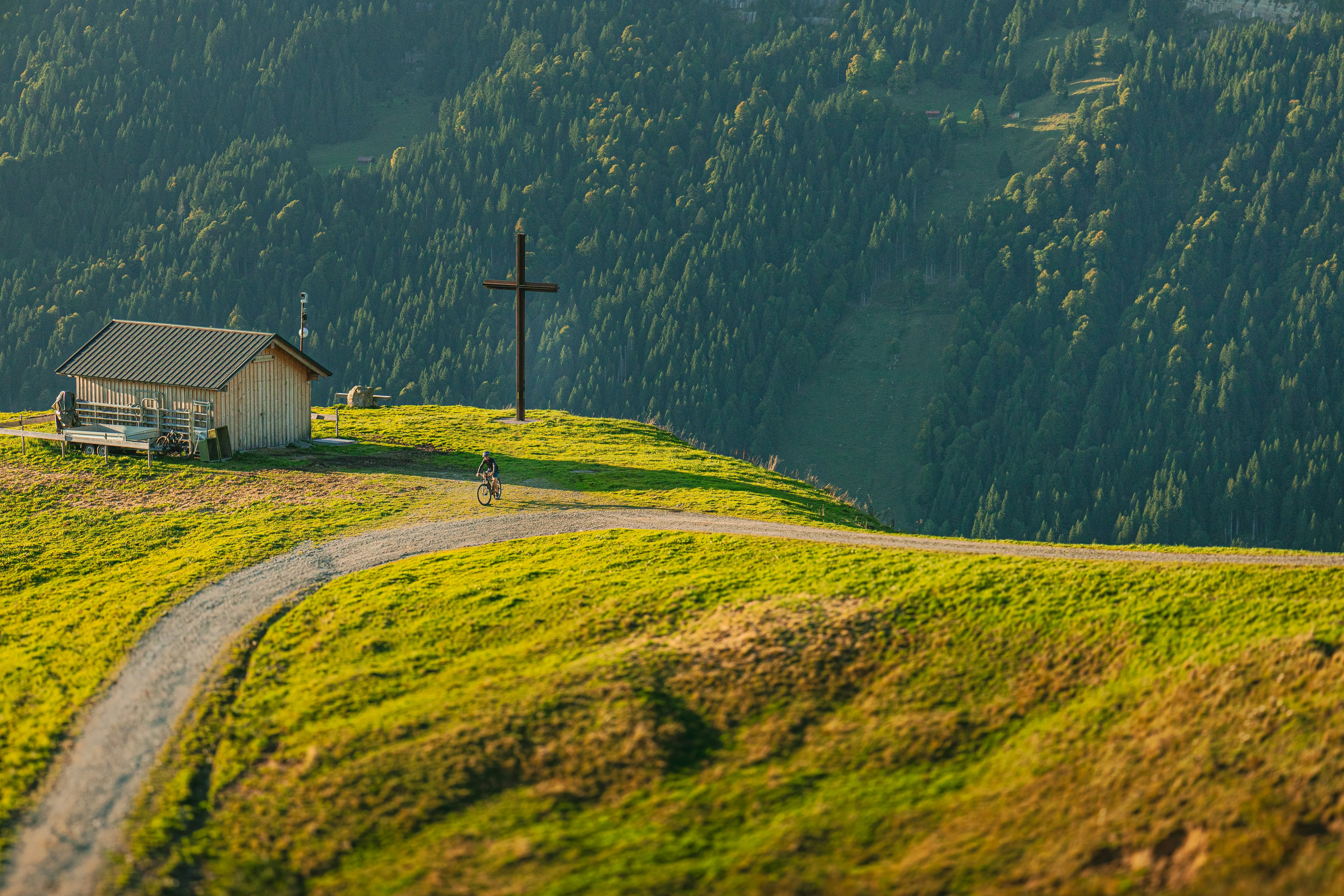 A cyclist traversing a winding path on a lush hillside, with a rustic cabin and a towering cross in the background. The scene captures the tranquility of mountain landscapes.