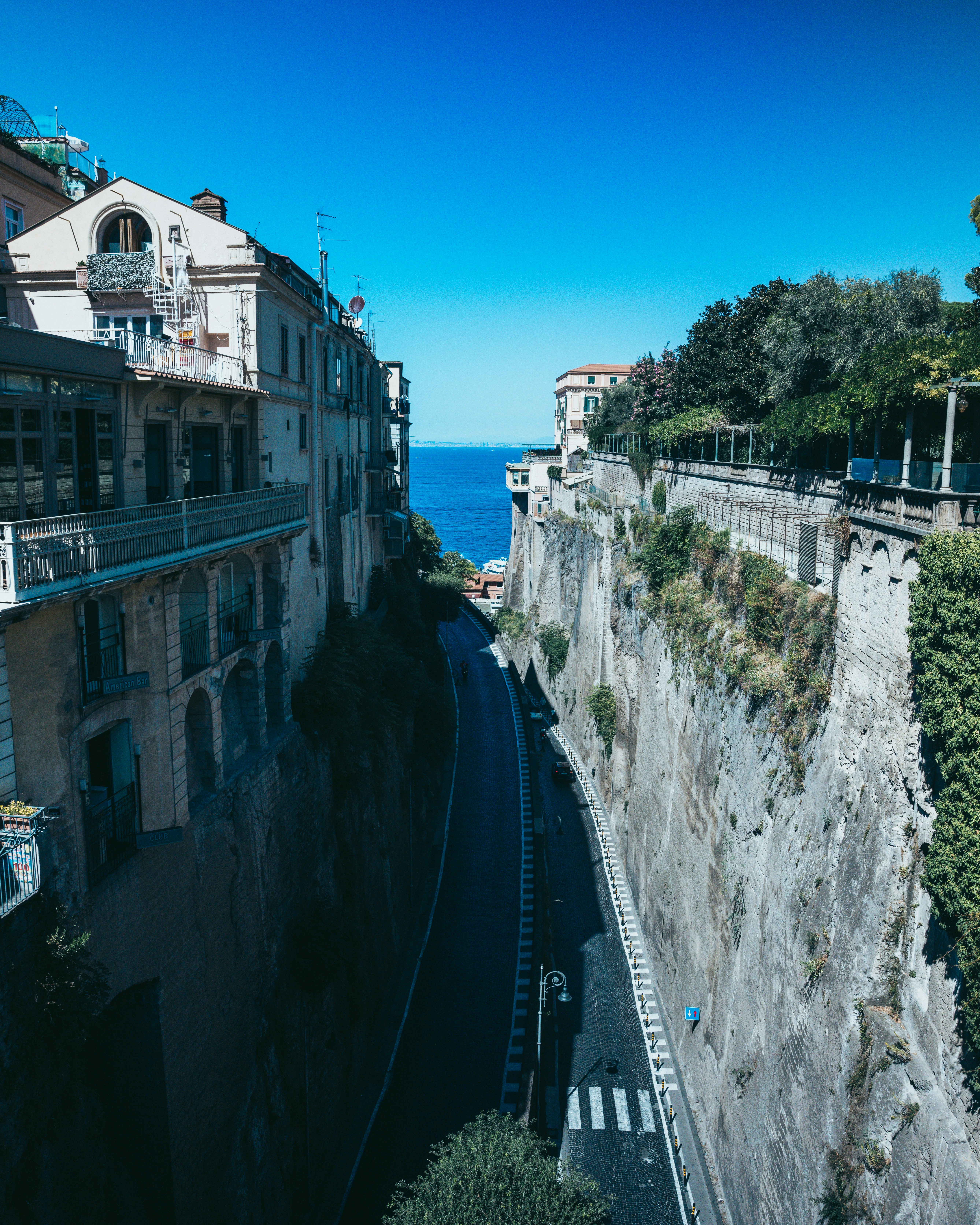 Narrow road flanked by towering cliffs and charming buildings, leading to the tranquil sea beyond.