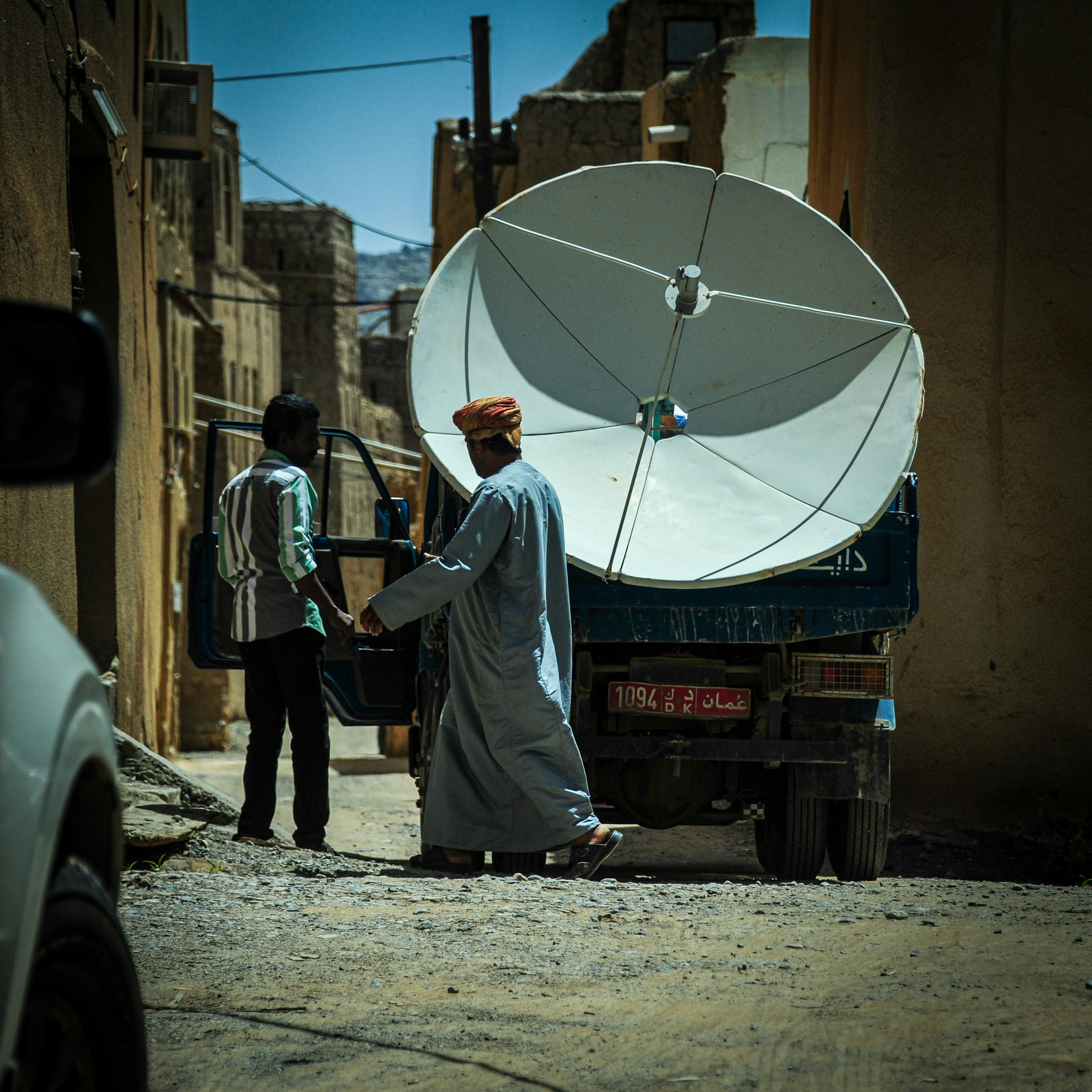 Two men interact while unloading a large satellite dish from a vehicle in a narrow alleyway, showcasing local life and technology.
