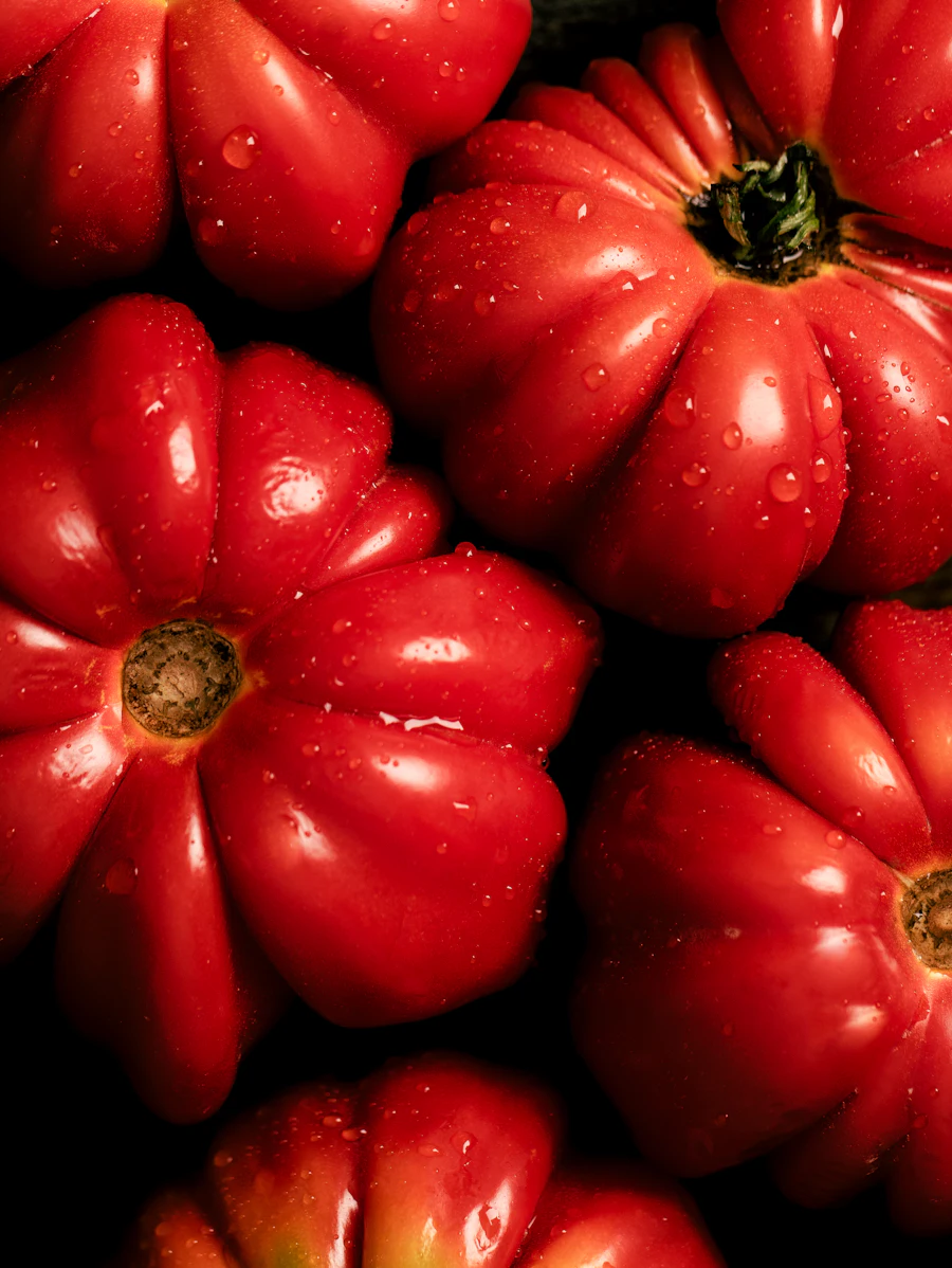 Close-up of clustered red heirloom tomatoes covered in water droplets against a dark background. Their ribbed shapes and glossy skins fill the frame.