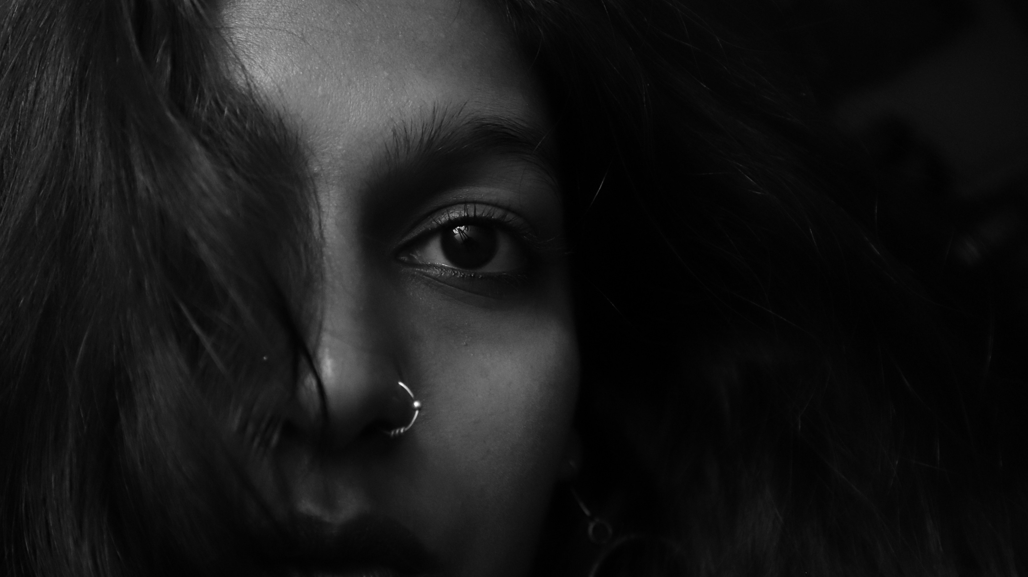 Close-up portrait of a person with flowing hair, emphasizing expressive eyes and a nose ring against a dark background.