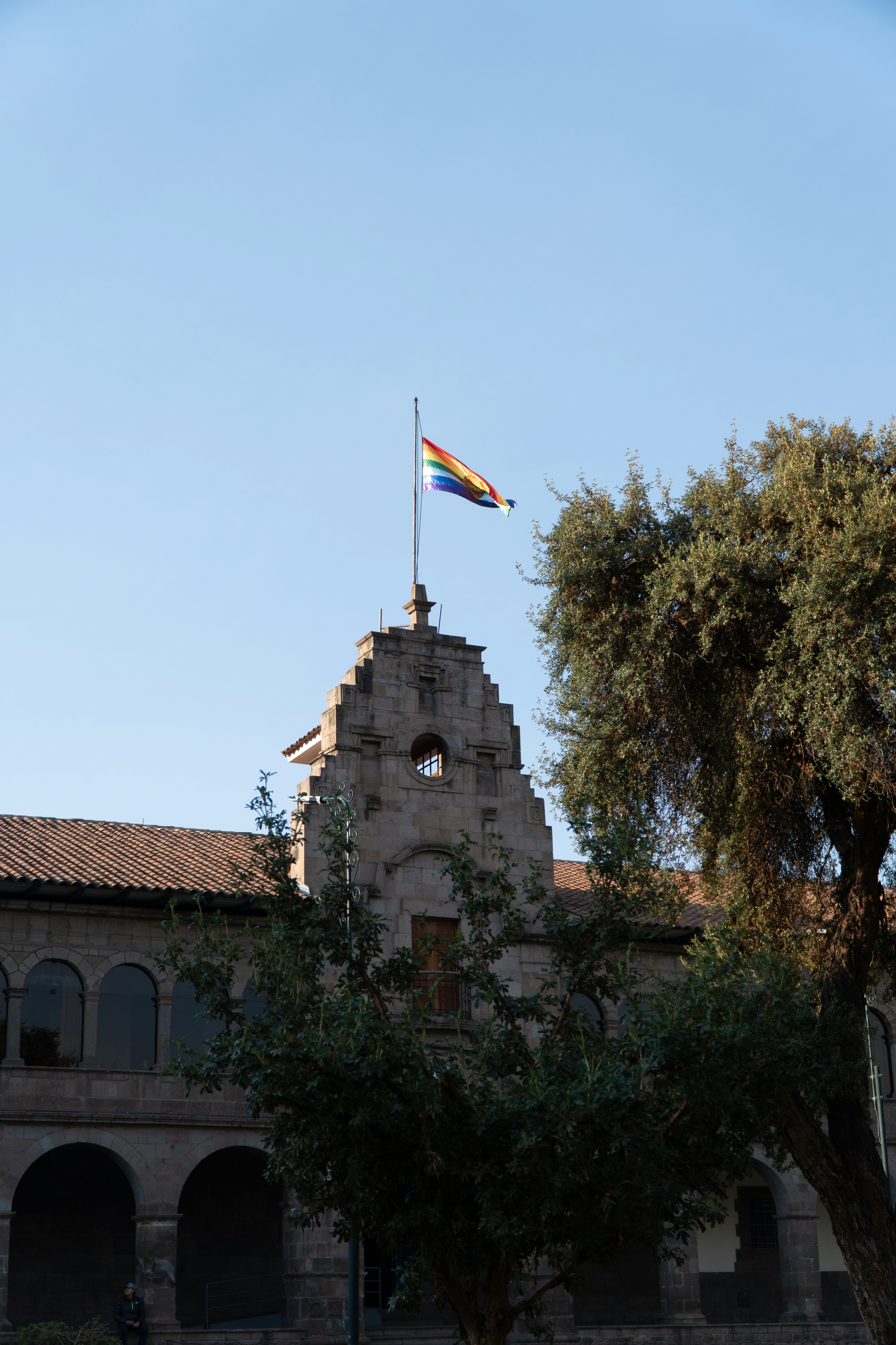 Pride flag proudly displayed atop a historic building, surrounded by lush trees under a clear sky.