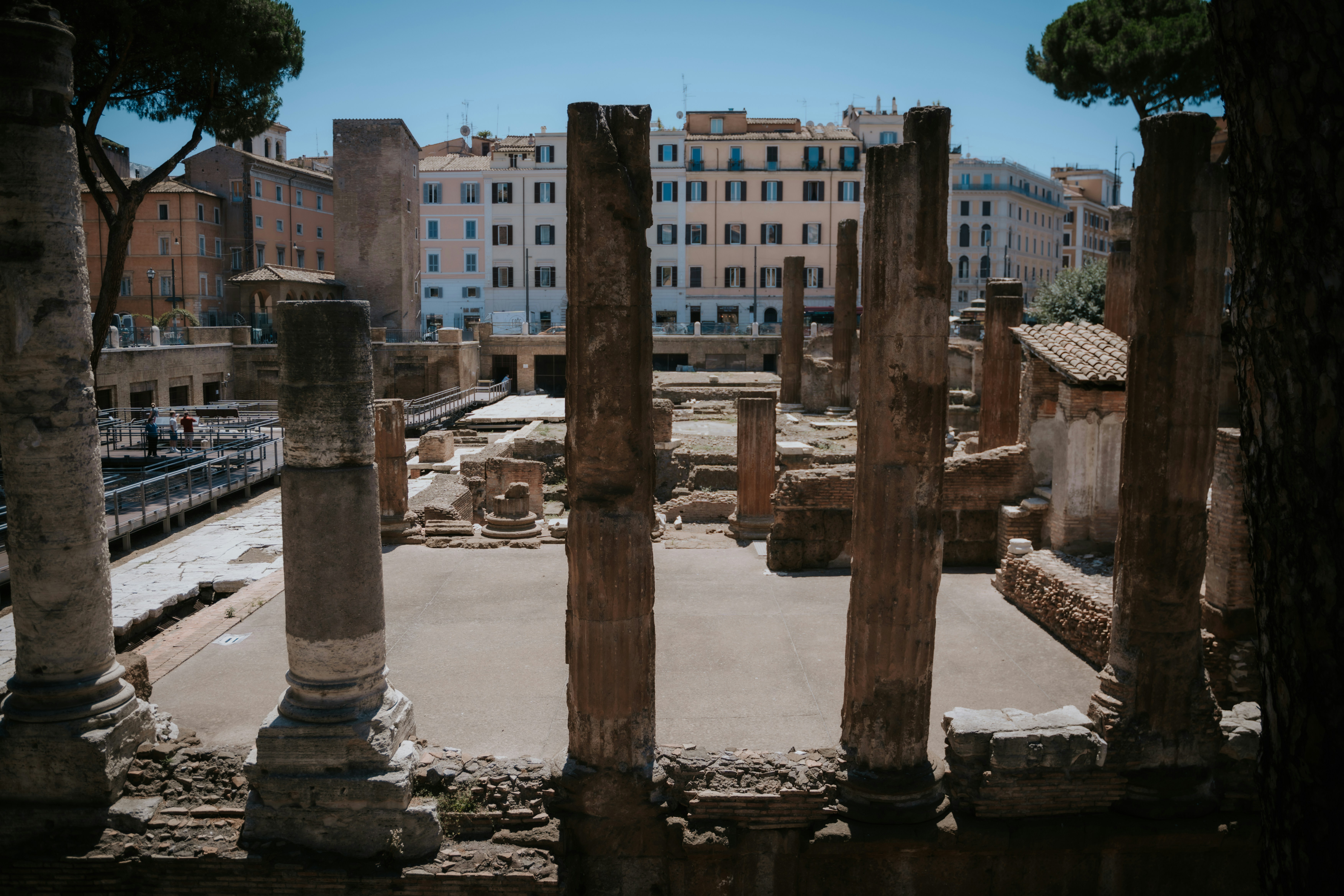 Ruins of ancient columns stand amidst the remnants of a historic site, framed by modern buildings in the background. A blend of history and contemporary life.