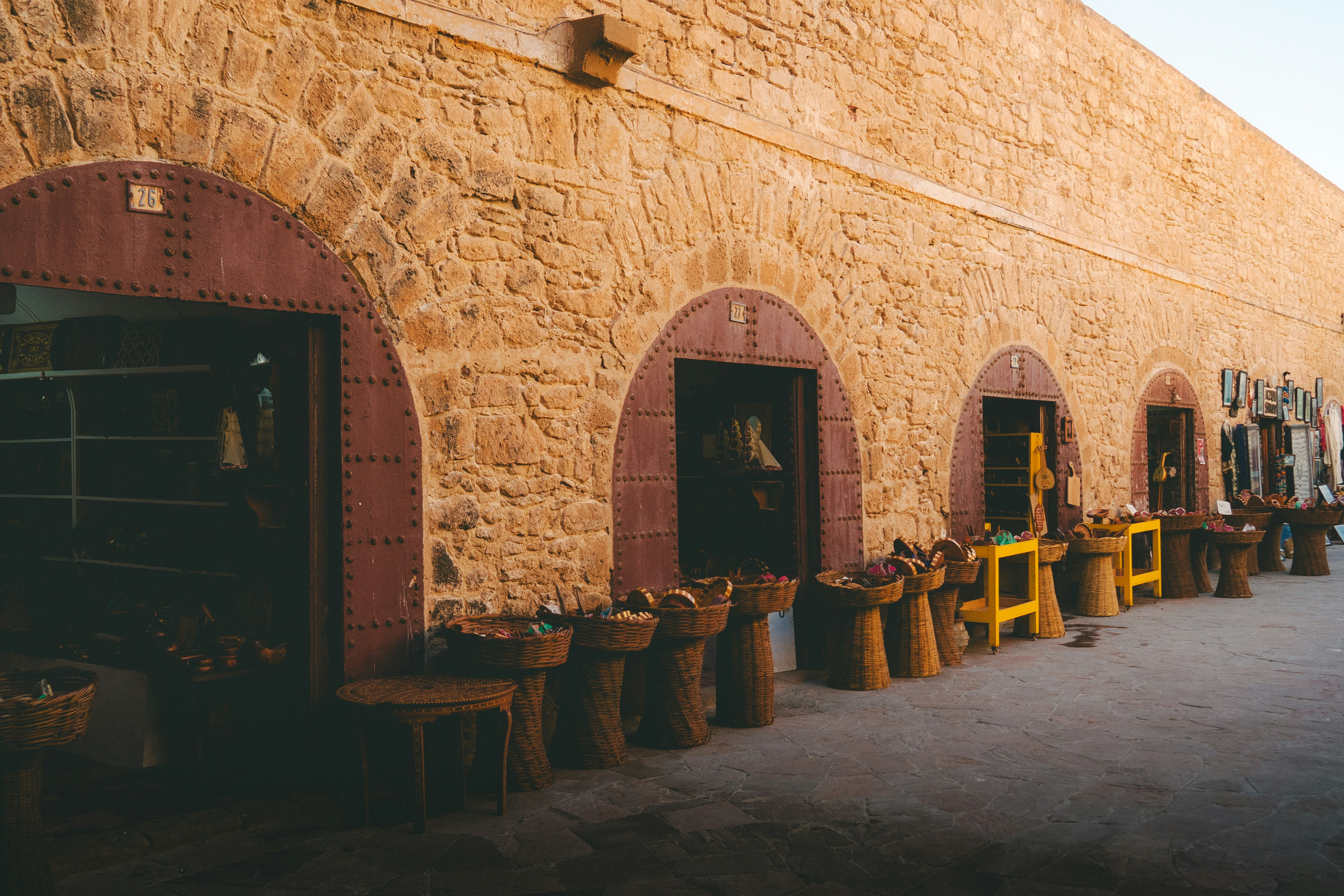 Vibrant market scene featuring arched doorways with woven baskets and colorful displays. The rustic stone wall adds character to the bustling atmosphere.