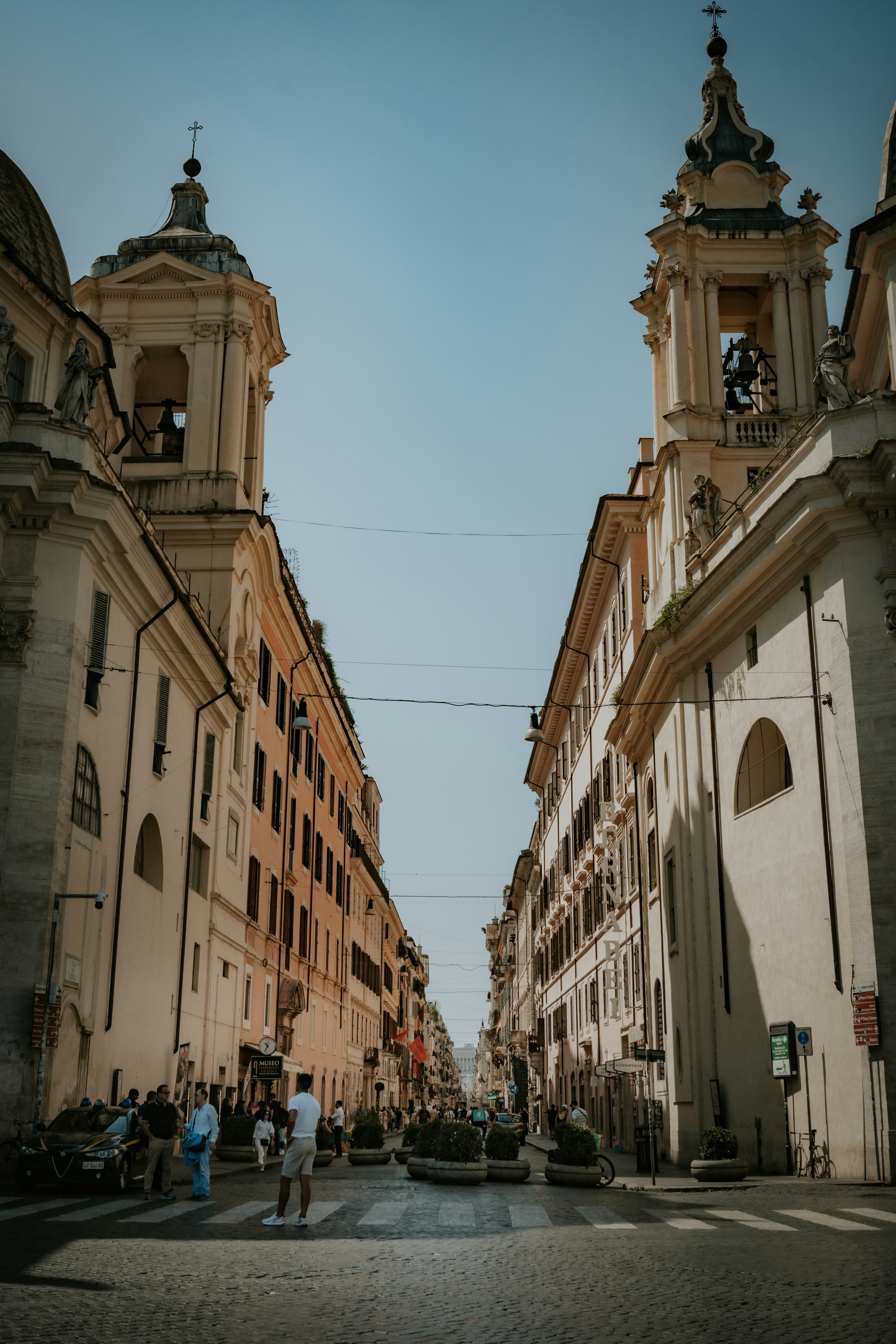 Narrow street in Rome framed by historical buildings, with pedestrians and greenery lining the path.