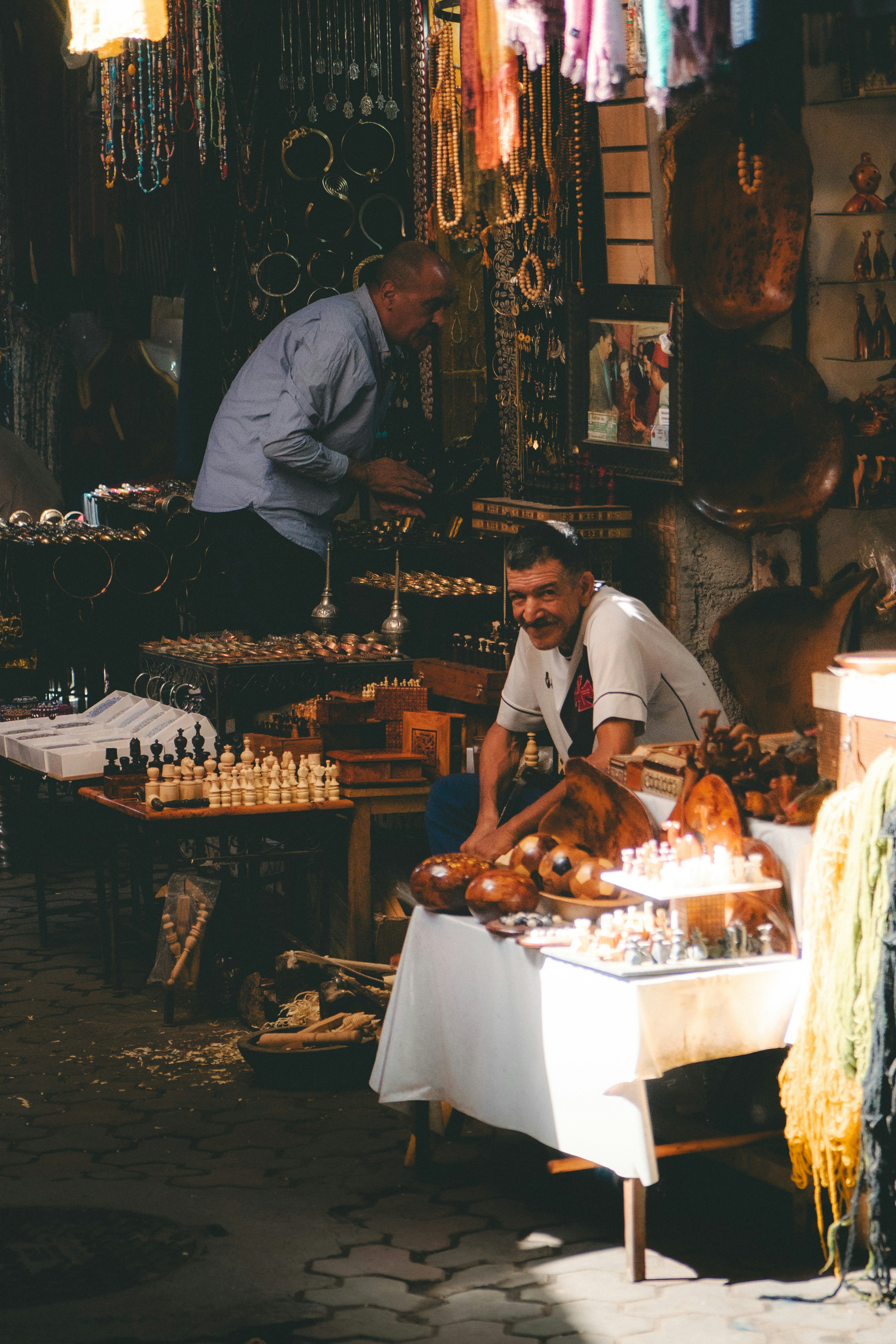 A bustling market scene featuring a vendor surrounded by handcrafted goods, including chess sets and jewelry, with another seller in the background. 
