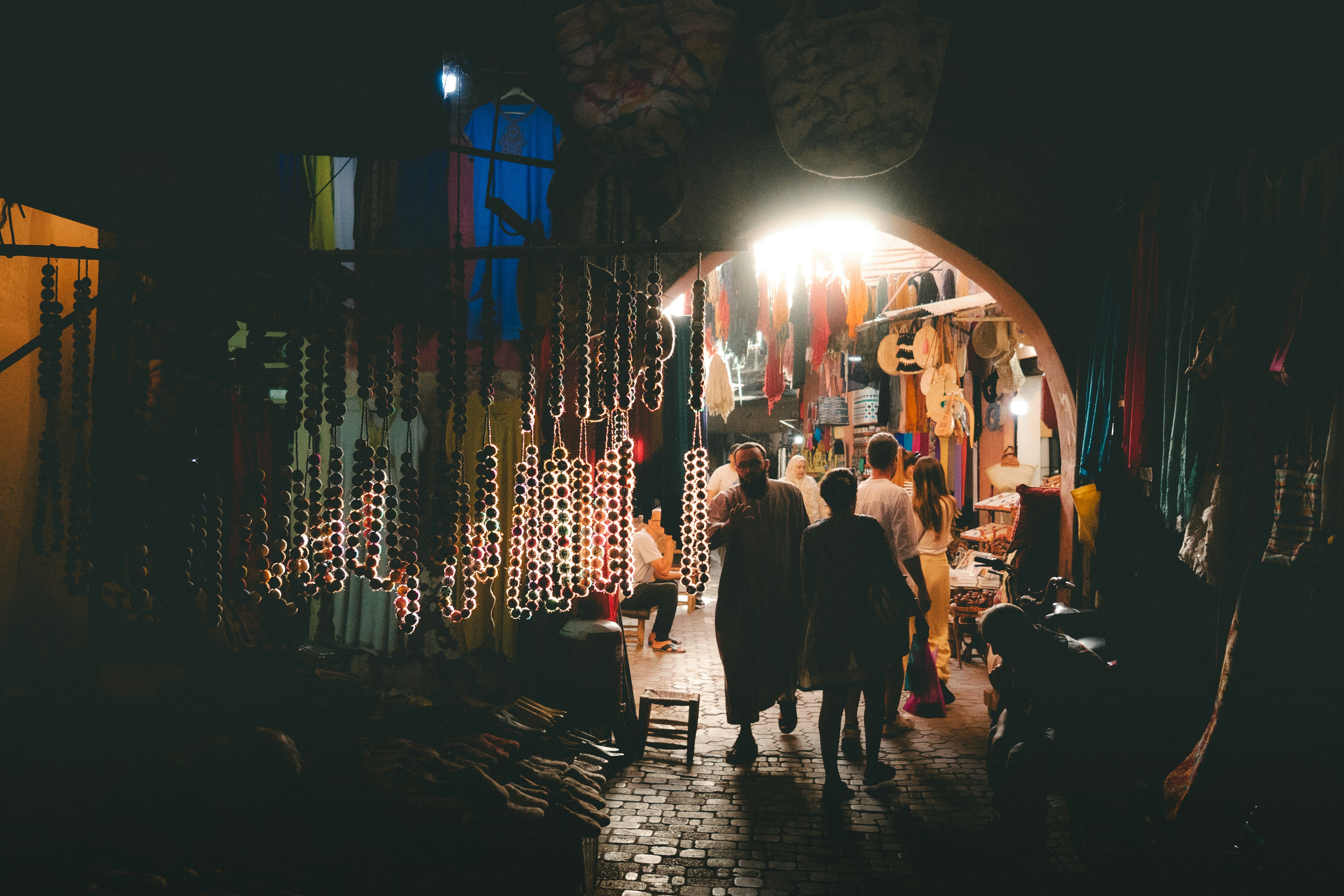 Vibrant market scene illuminated by warm lights, showcasing colorful textiles and bustling shoppers in a narrow alleyway.