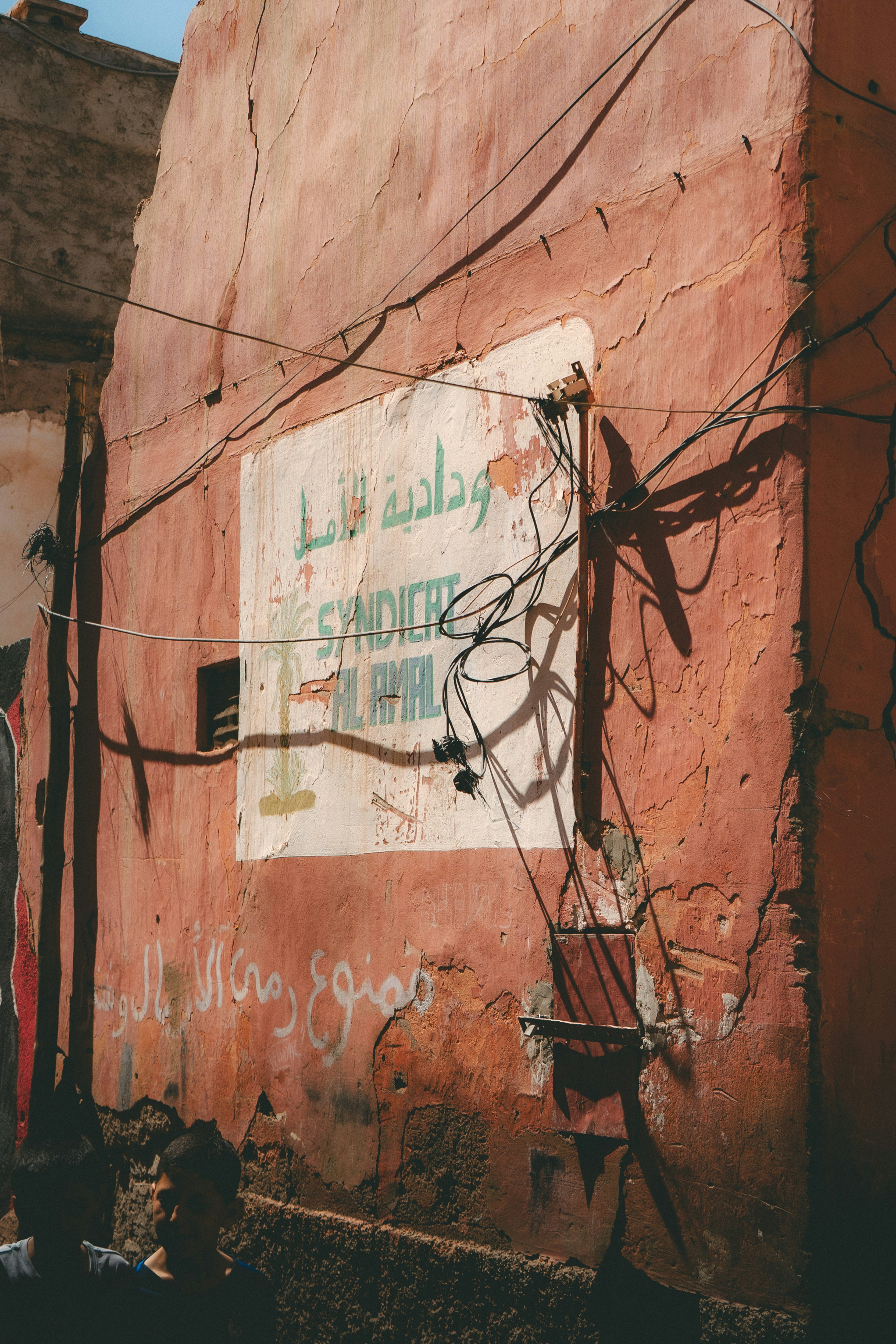 Wired Shadows: A Green Sign Adrift on a Weathered Pink Facade
