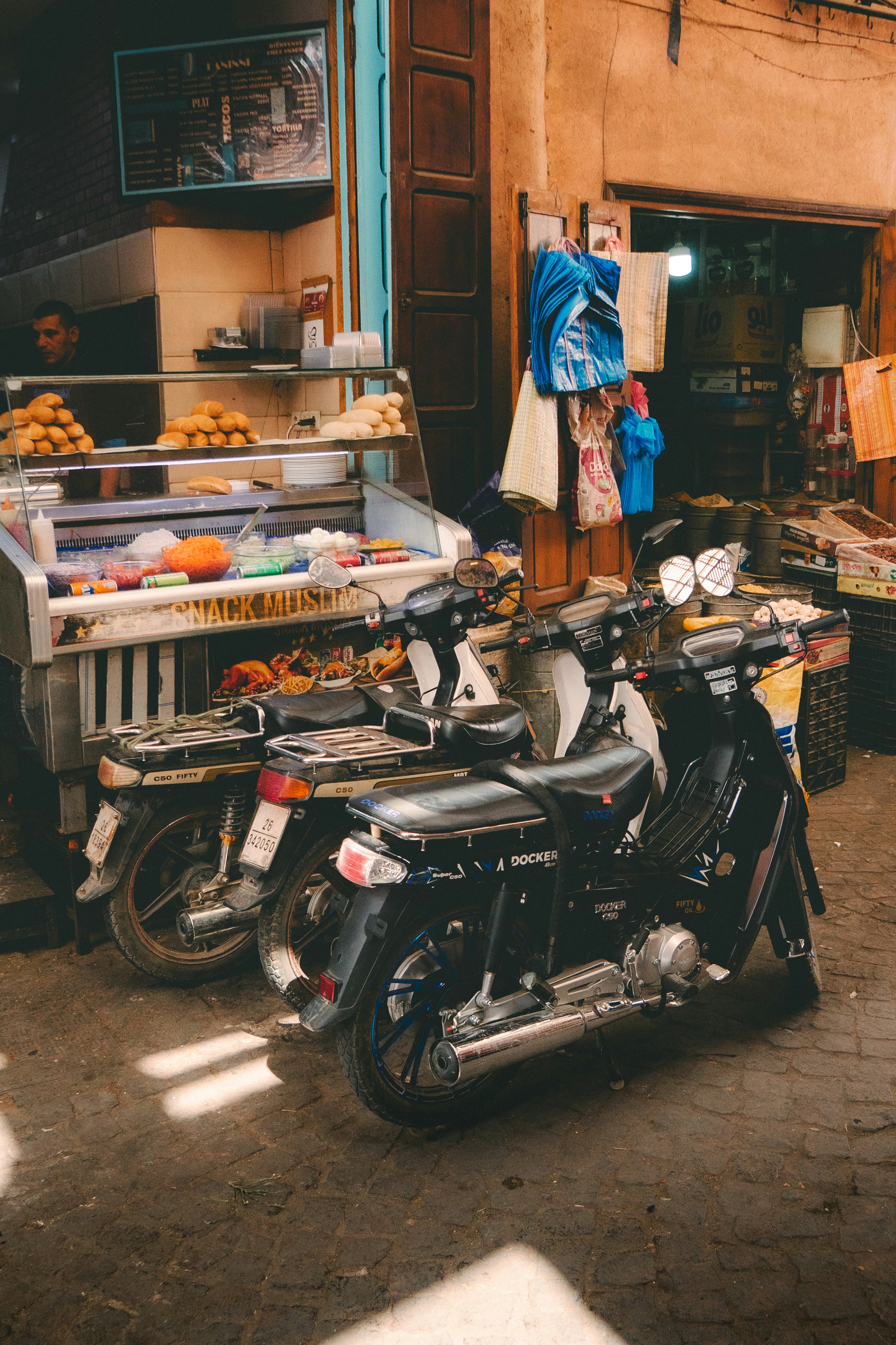 Three parked motorbikes in a bustling market scene, surrounded by food stalls and vibrant colors. The atmosphere captures the essence of daily life in an urban setting.