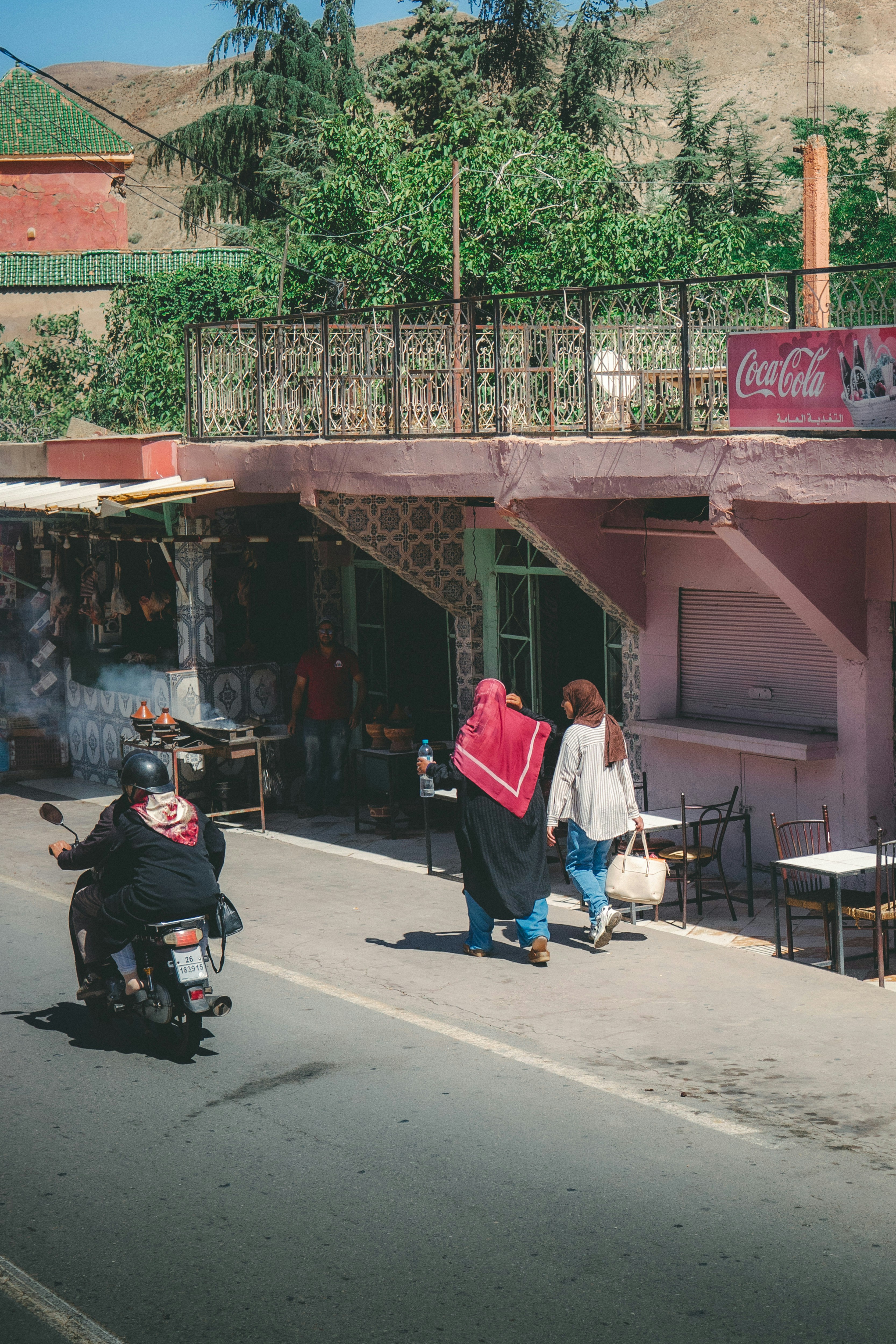 Two women walking along a bustling street in a lively marketplace, with a motorcycle passing by and shops in the background.