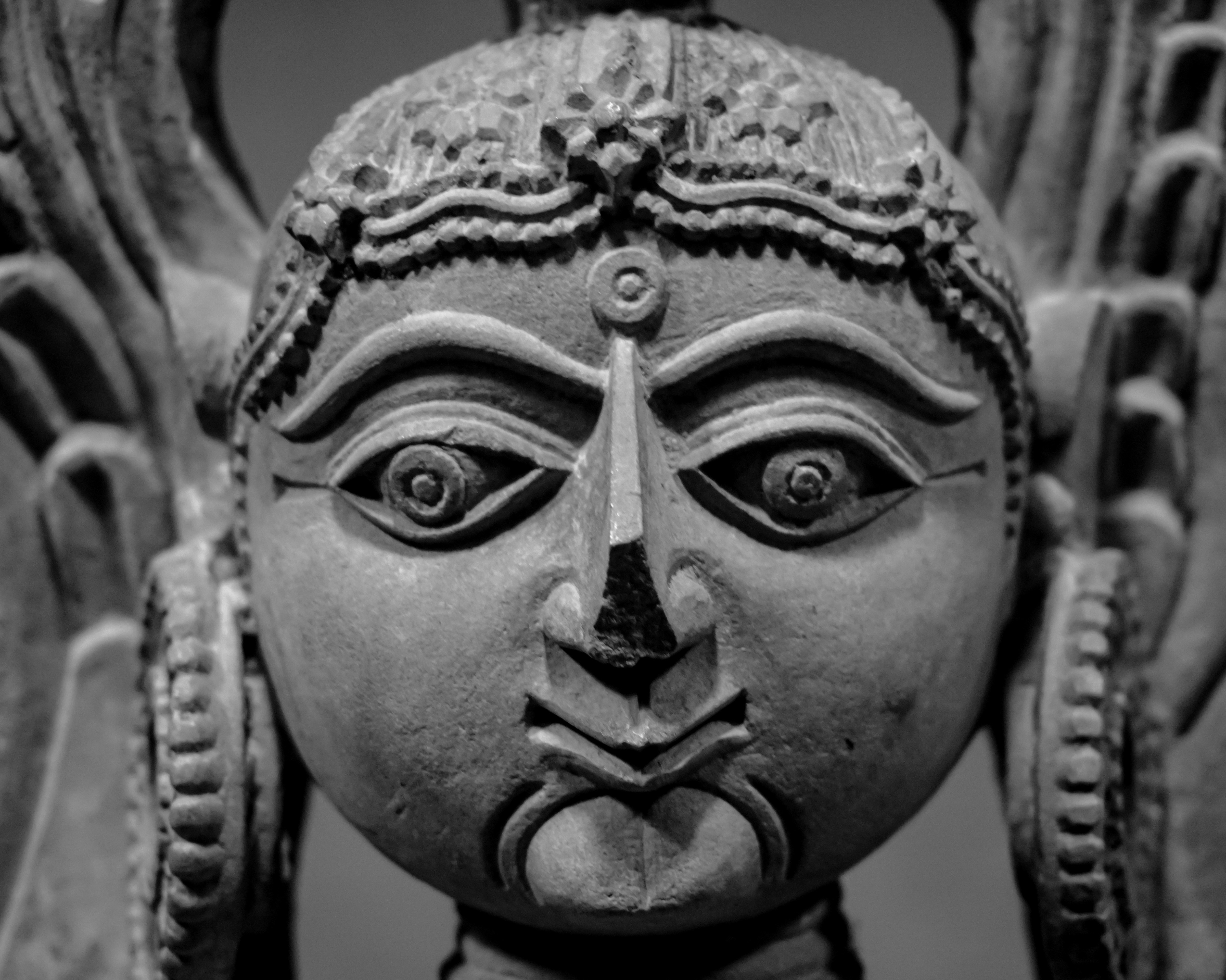 A close-up, black and white photograph of an ancient, winged stone sculpture's face. The sculpture features large, round eyes, an elongated nose, and a distinct mouth with a downturned expression. Intricate details adorn the forehead and what appears to be a headdress or crown, suggesting a deity or mythical figure. The texture of the stone is clearly visible, adding to its historical and artistic appeal. | Detailed stone carving of a mythical winged creature's face.
